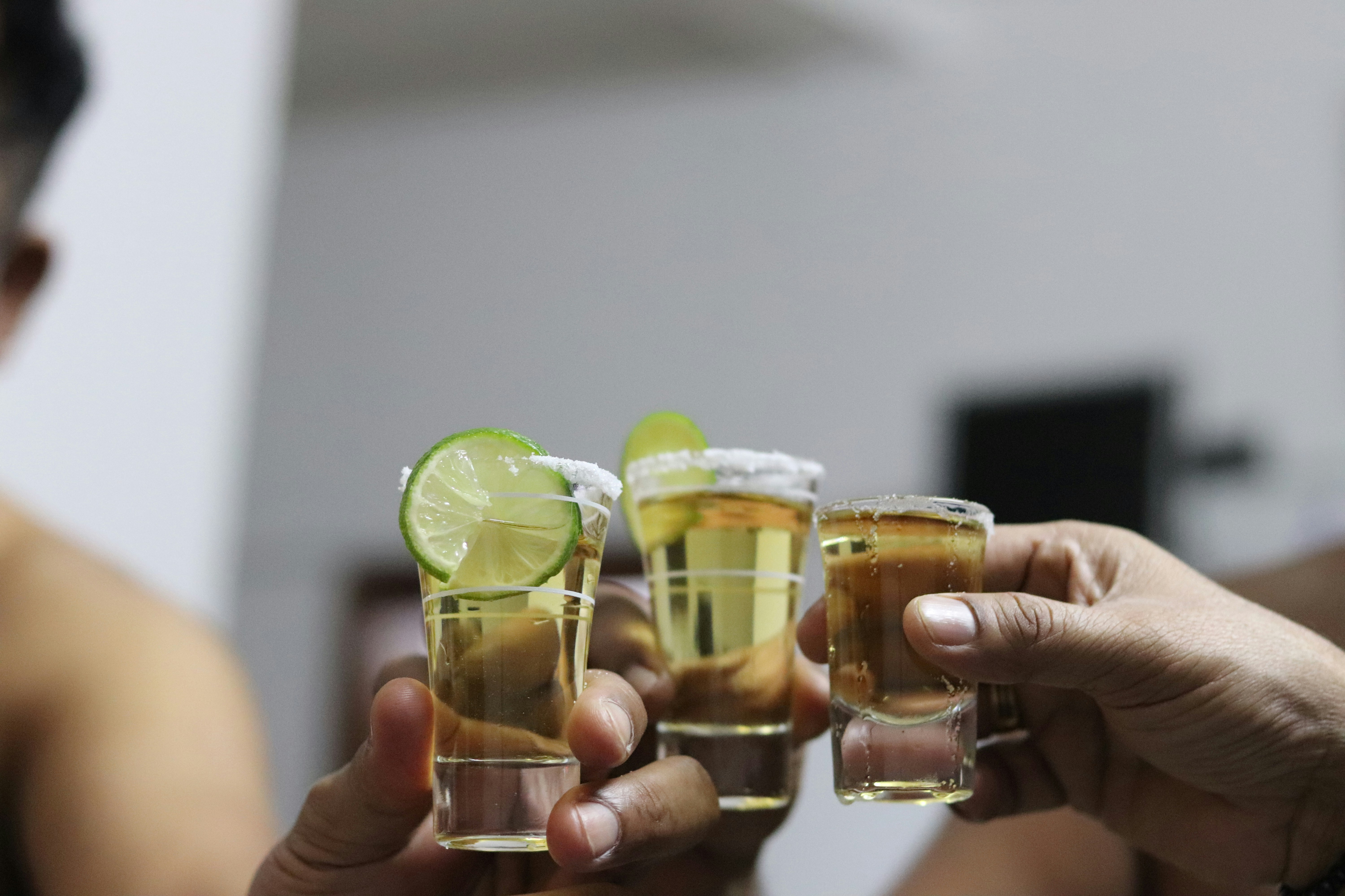 Three shot glasses filled with tequila, garnished with lime, held up in a celebratory toast. The background features a blurred party atmosphere.