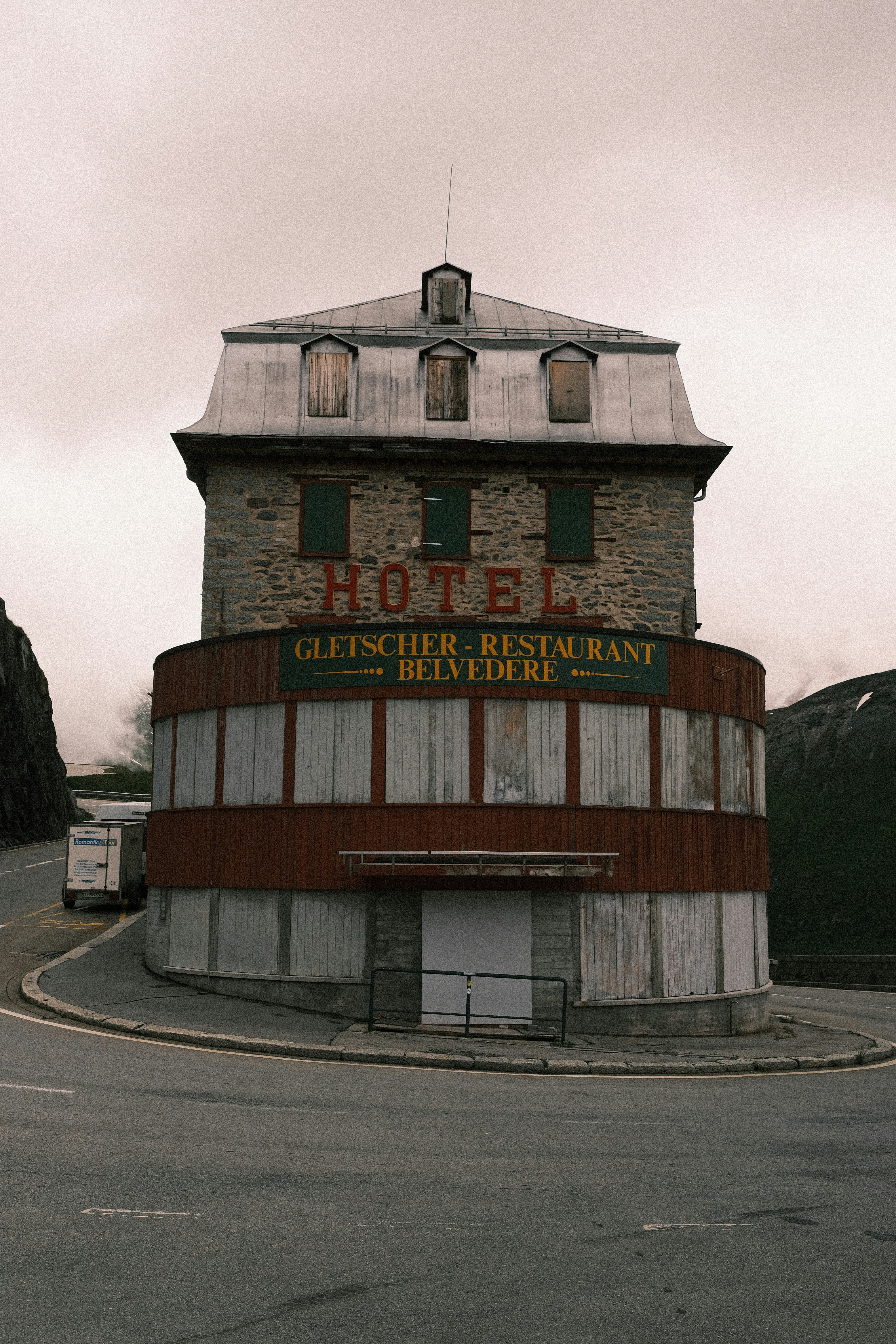 A vintage hotel structure with a rounded base and prominent signage, nestled against a mountainous backdrop. The building showcases a blend of stone and wood, reflecting its rich history.