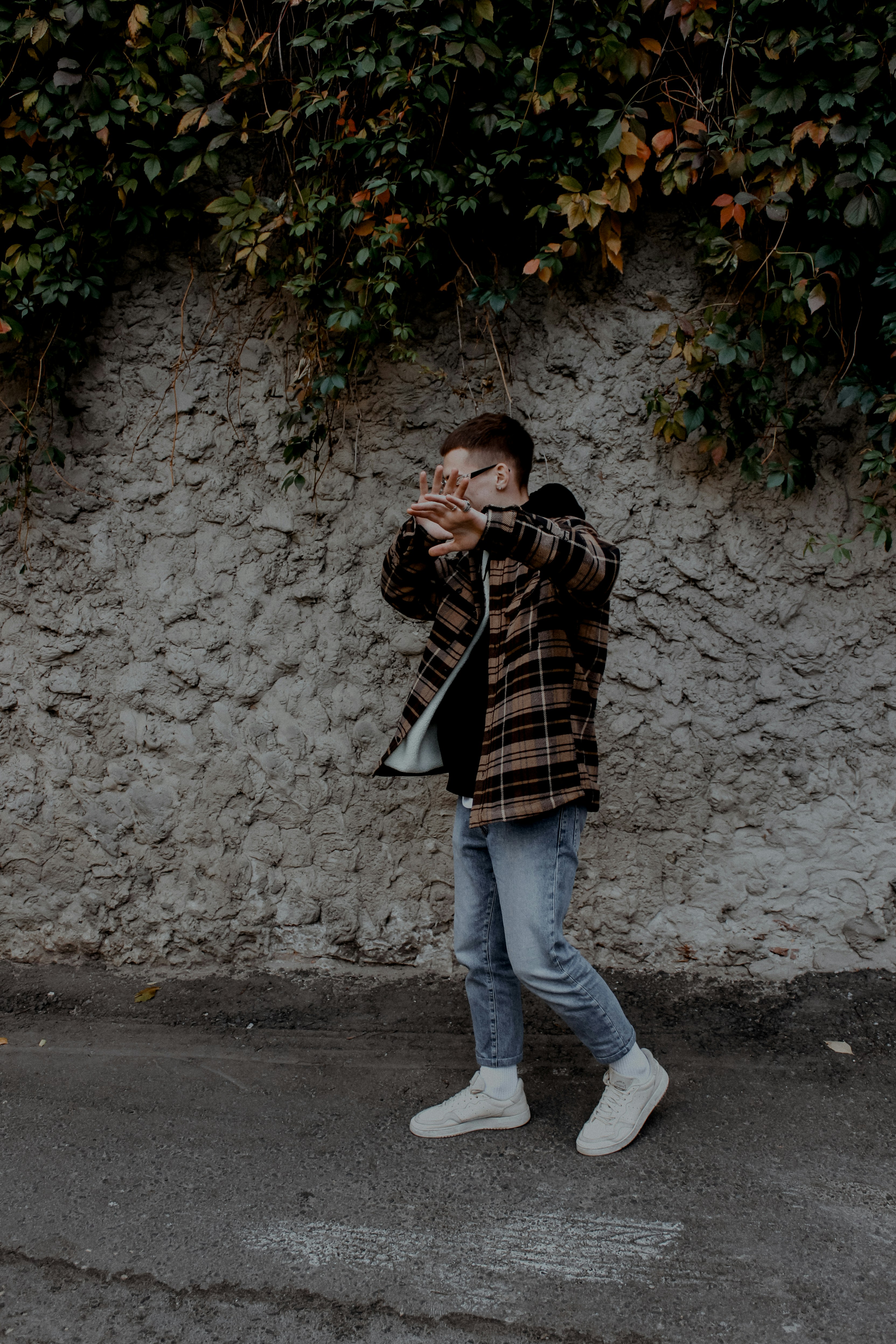 Young man in plaid jacket poses against a textured wall adorned with greenery, capturing an urban vibe. His hand gestures suggest a moment of contemplation.