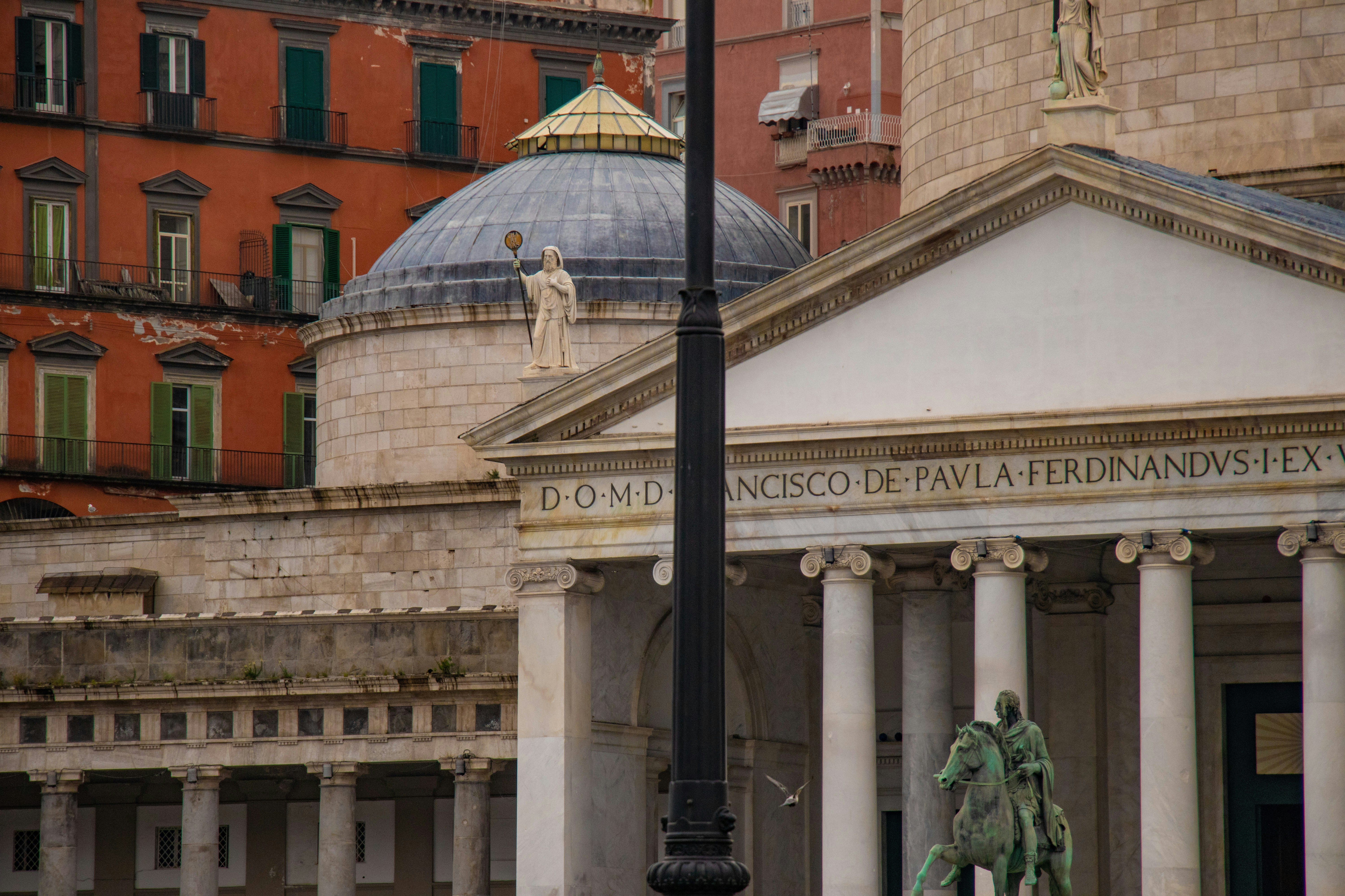 Detailed view of classical columns and a dome with a statue set against vibrant urban buildings.