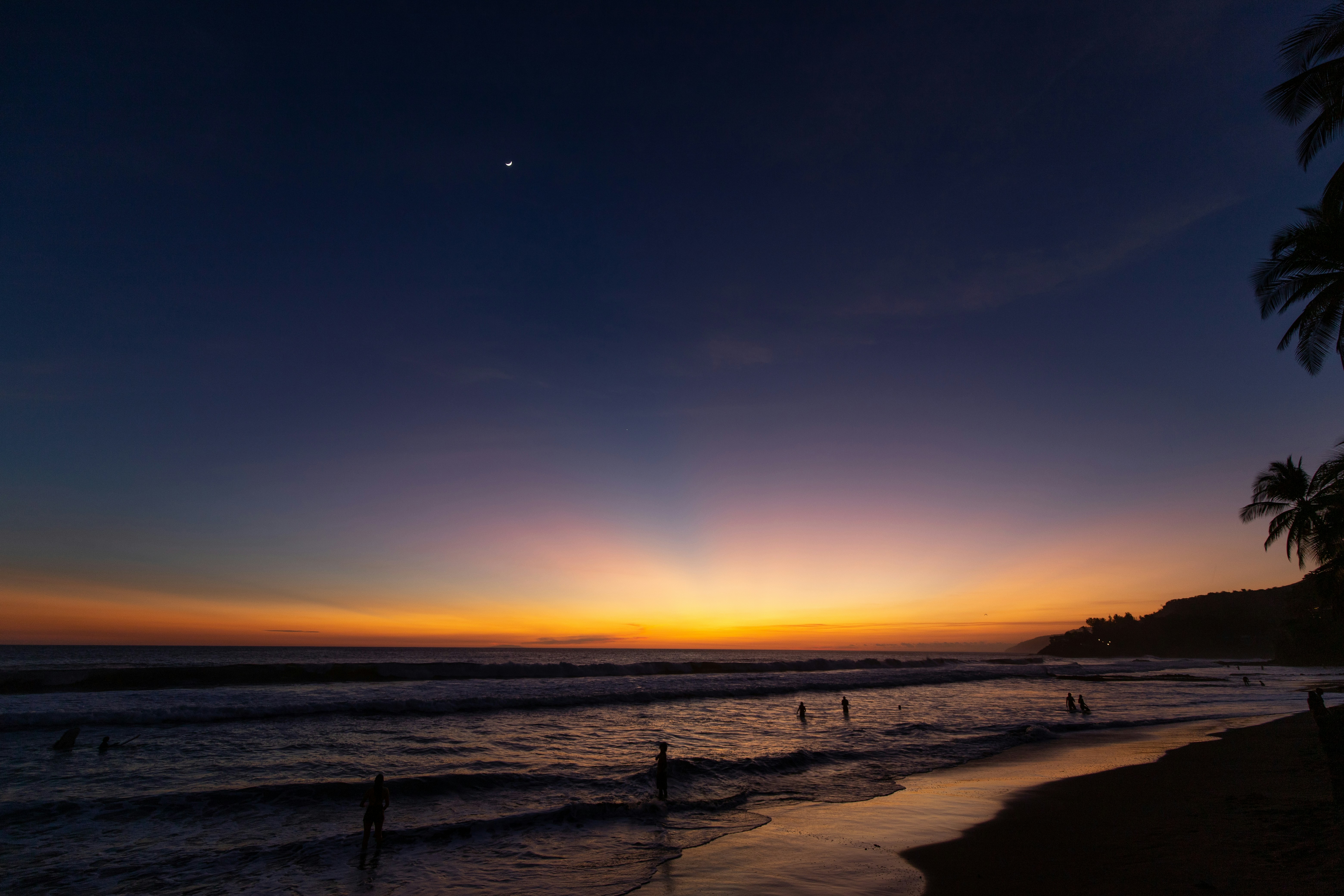 Vibrant sunset casting hues over a tranquil beach with silhouettes of people enjoying the water. The moon is faintly visible in the twilight sky.