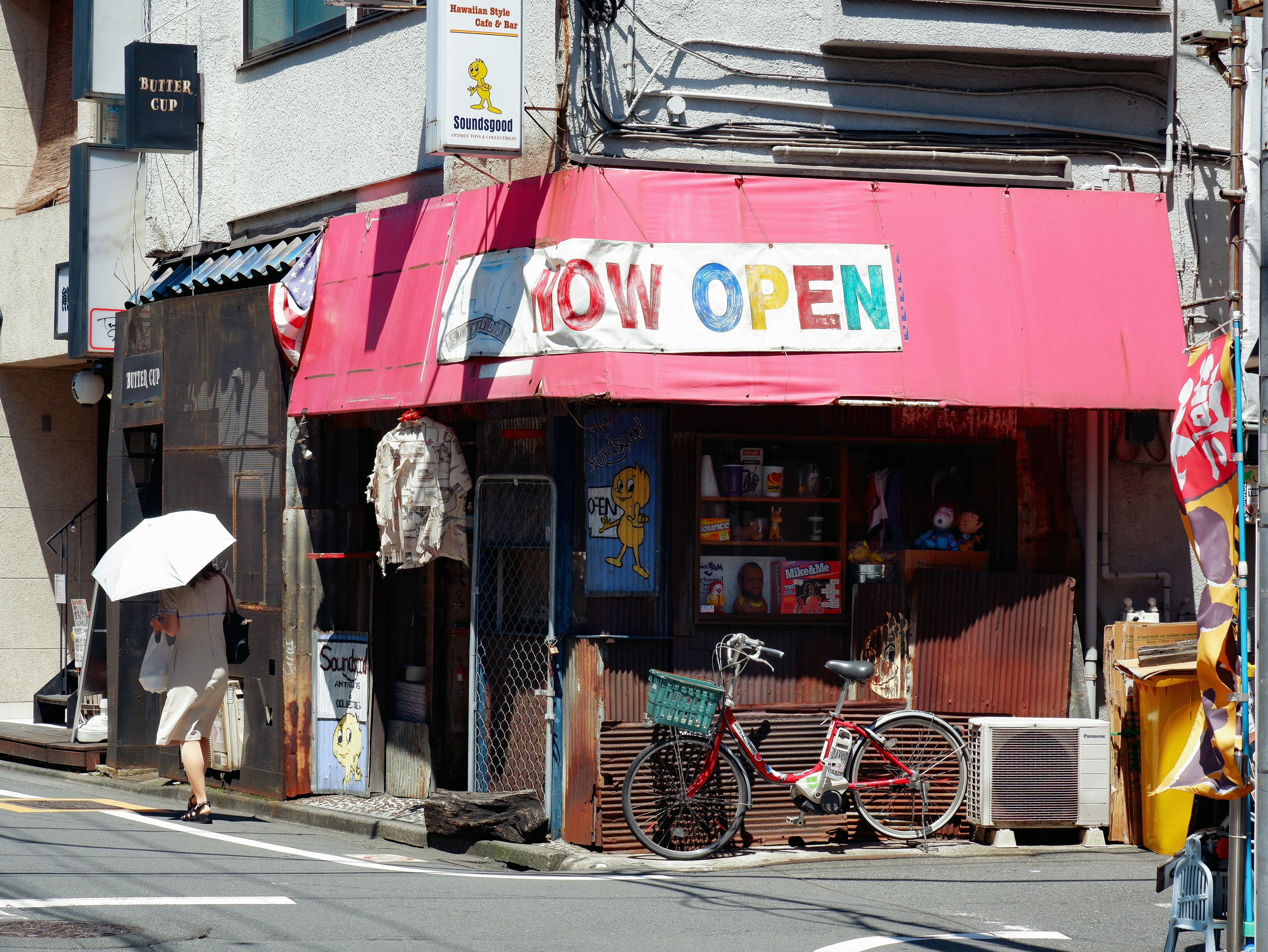 Quaint storefront with a bright pink awning displaying 'NOW OPEN,' featuring a bicycle and a passerby with an umbrella. The scene captures local life and character.