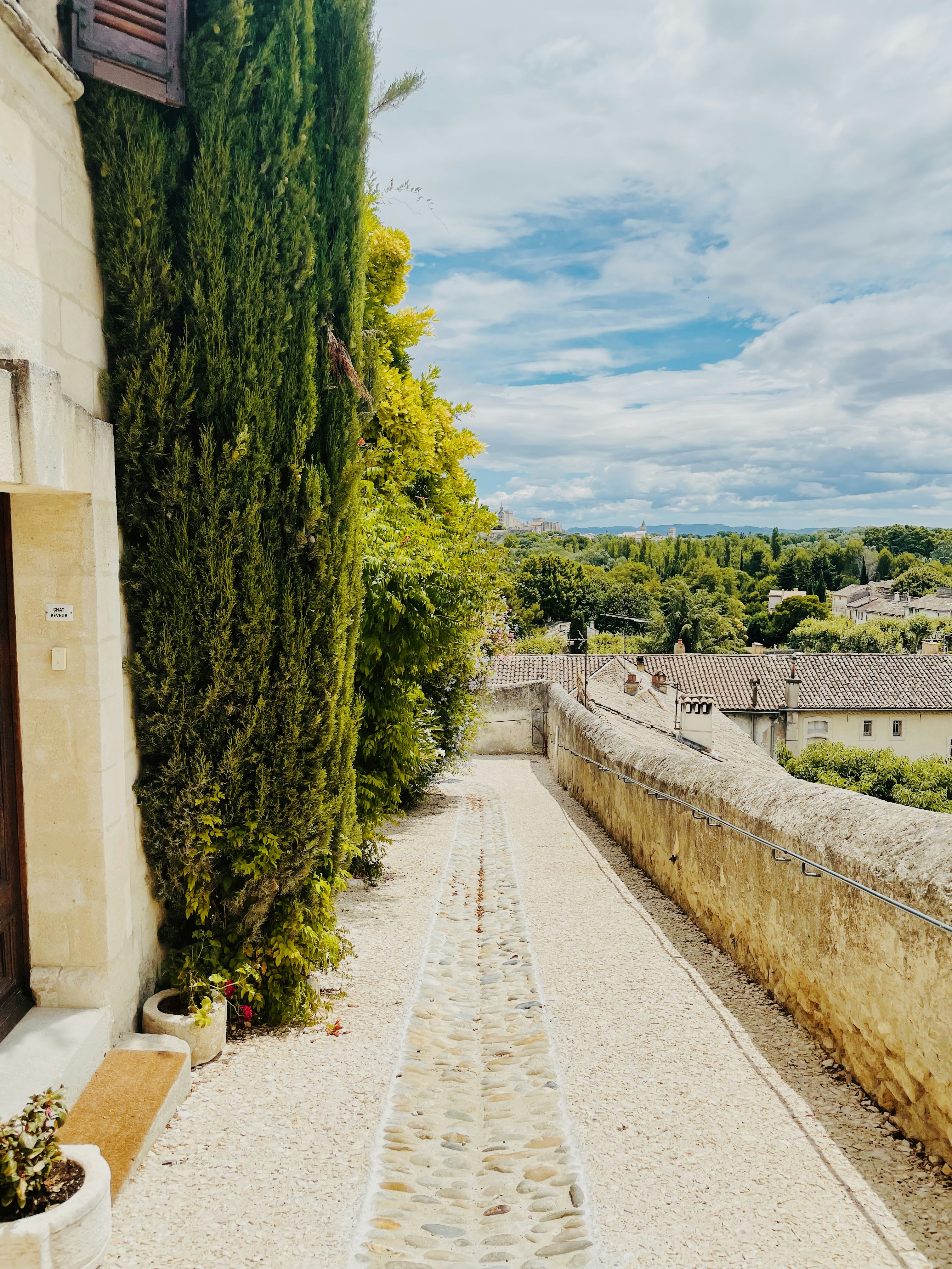Narrow cobblestone pathway flanked by lush greenery and historic stone walls under a partly cloudy sky.
