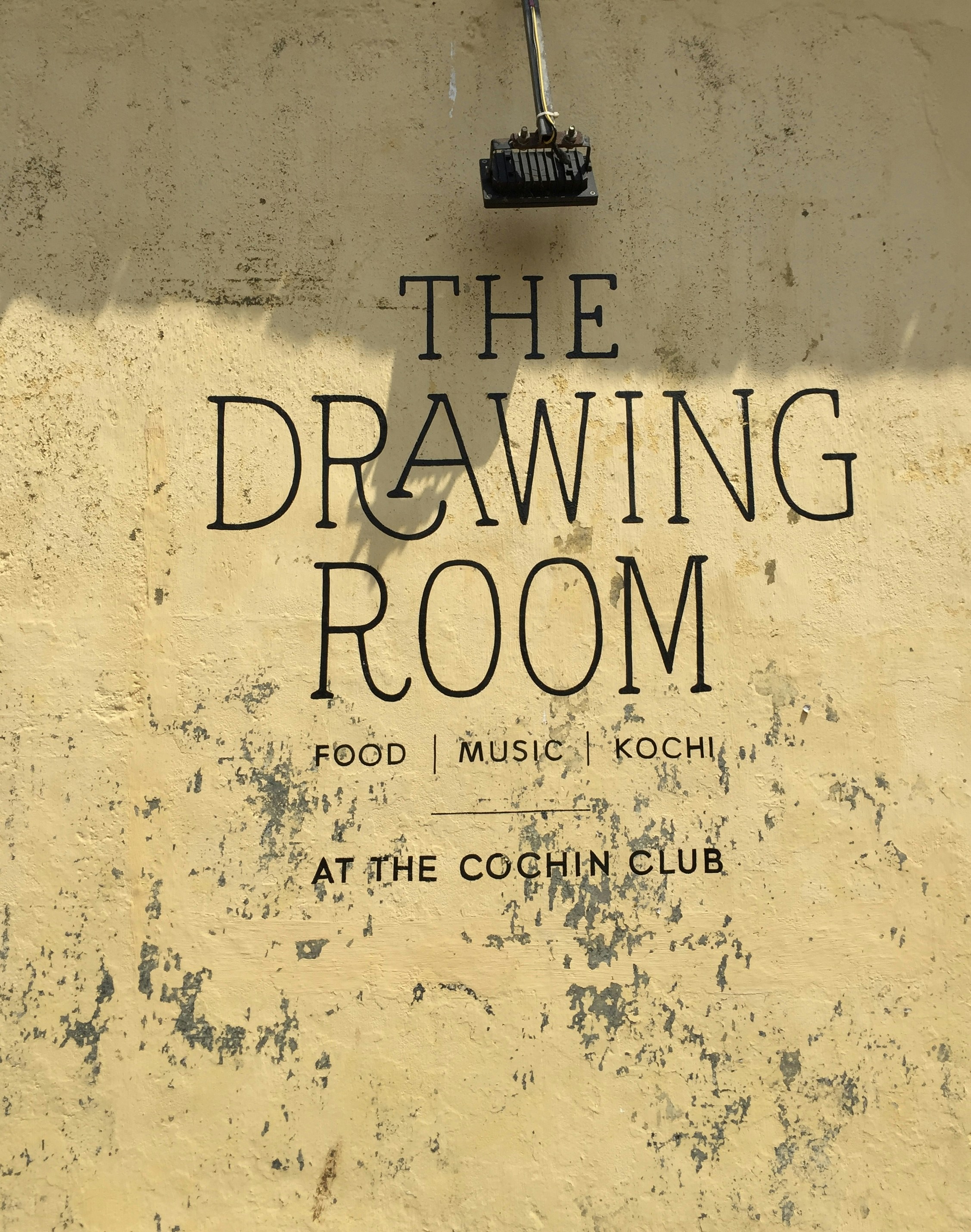 Stylish wall signage for The Drawing Room, highlighting its offerings of food and music in Kochi. The textured yellow background adds warmth to the inviting atmosphere.