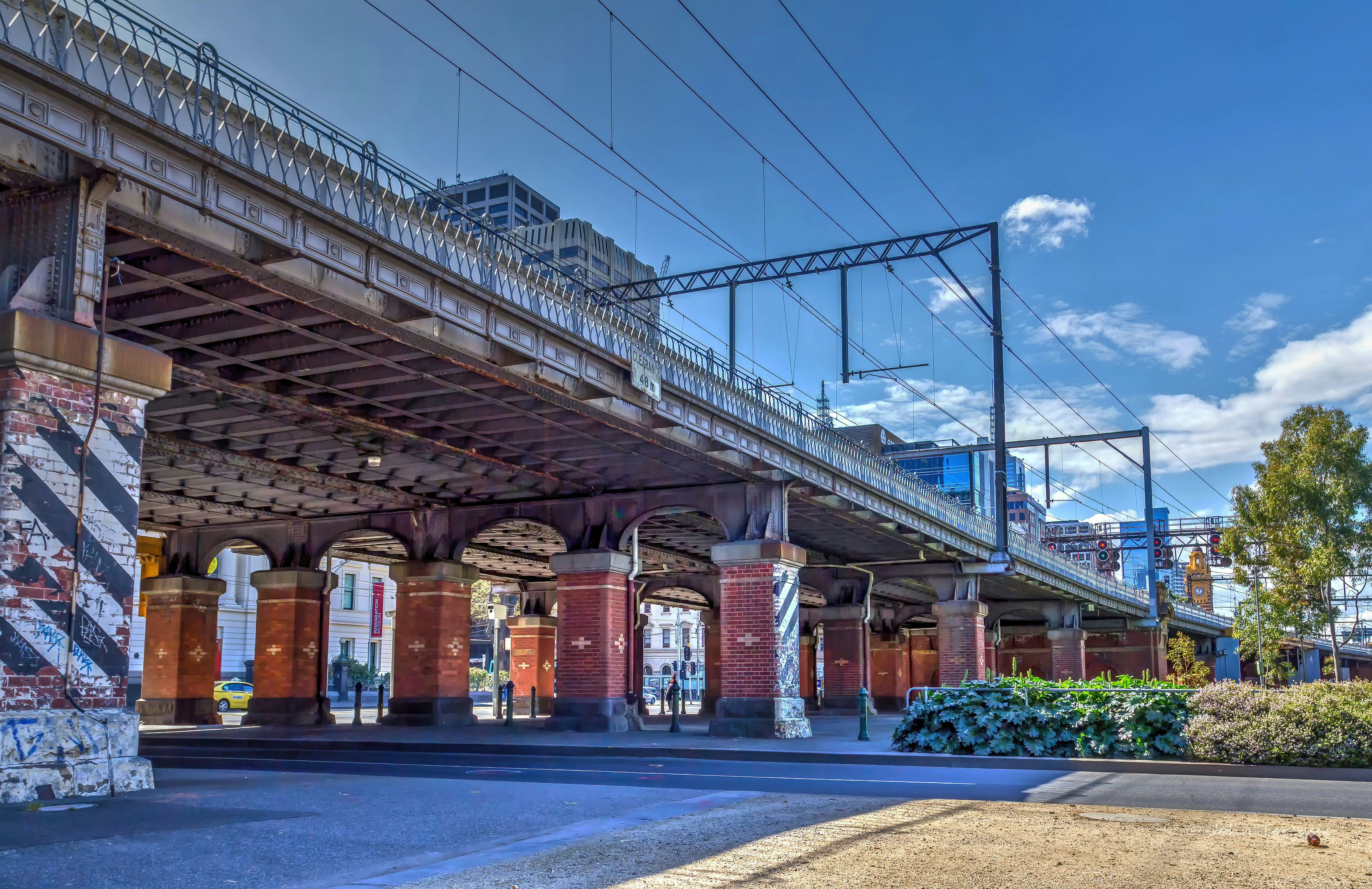 Vibrant urban landscape featuring a railway overpass supported by intricately designed pillars, juxtaposed with greenery and city buildings in the background.