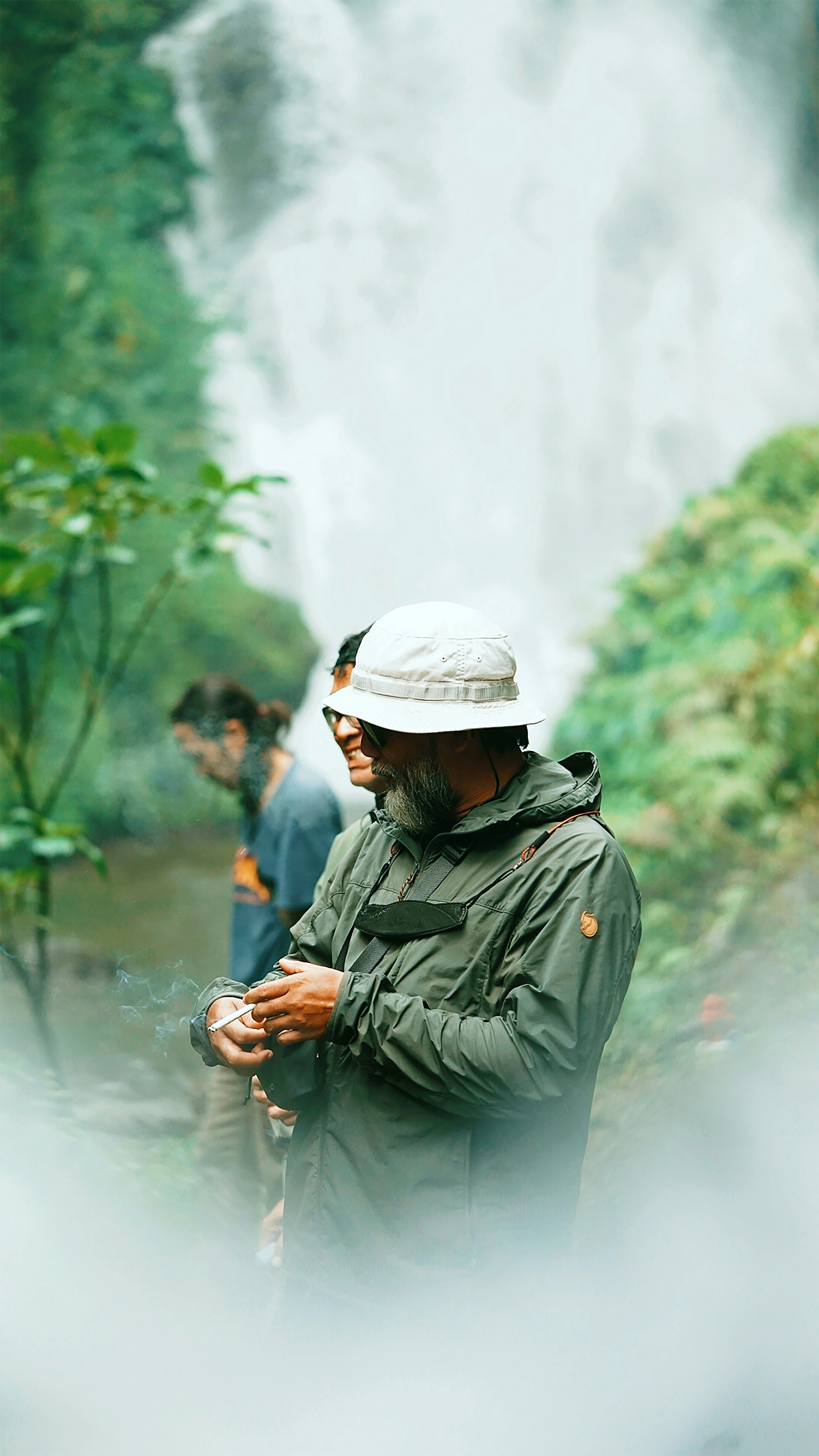 A man in a green jacket and white bucket hat stands by a waterfall, surrounded by lush greenery and soft mist. His contemplative pose suggests a moment of reflection.