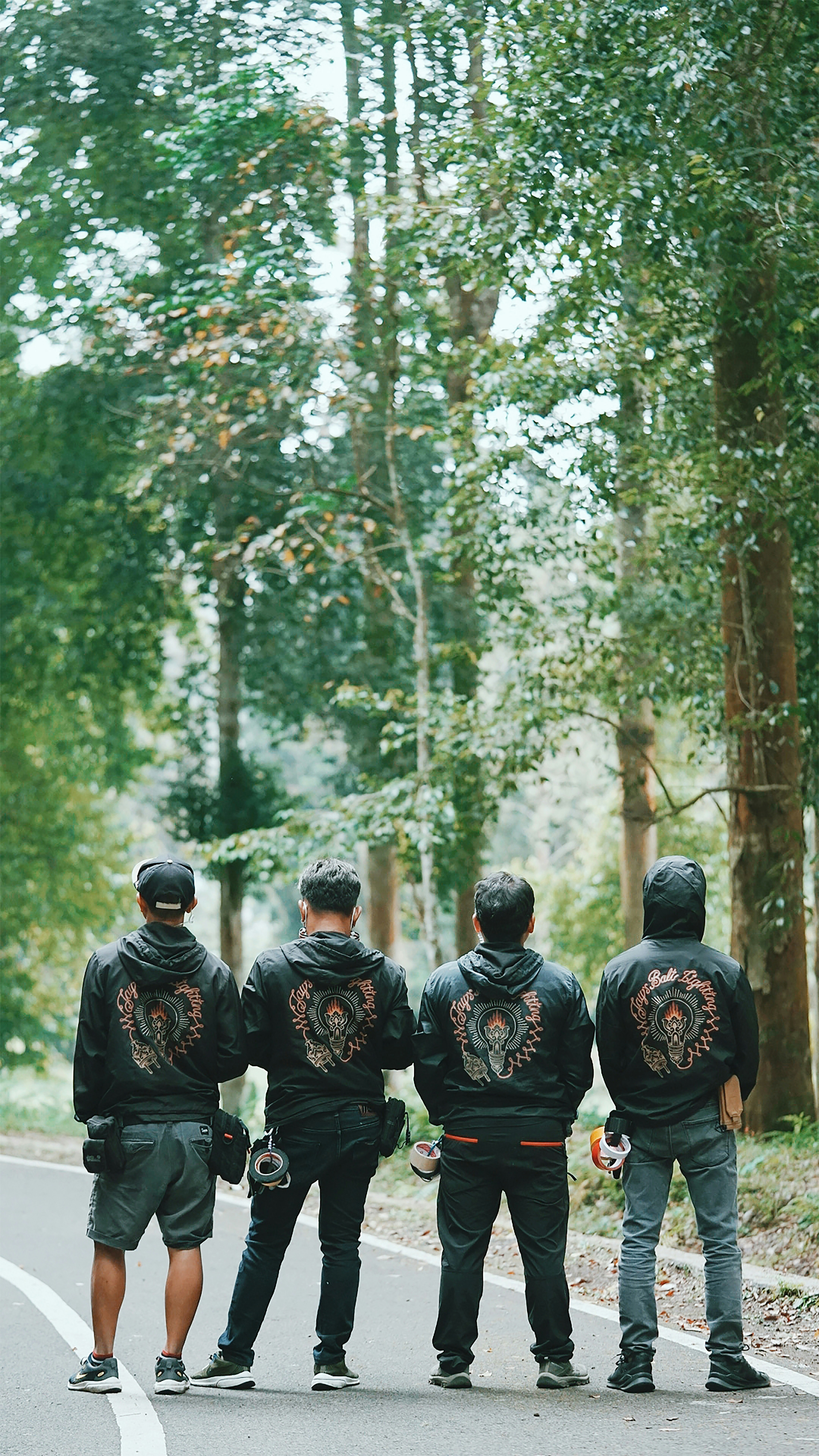 Four individuals in matching jackets stand on a winding road, surrounded by lush greenery, embodying camaraderie and adventure.