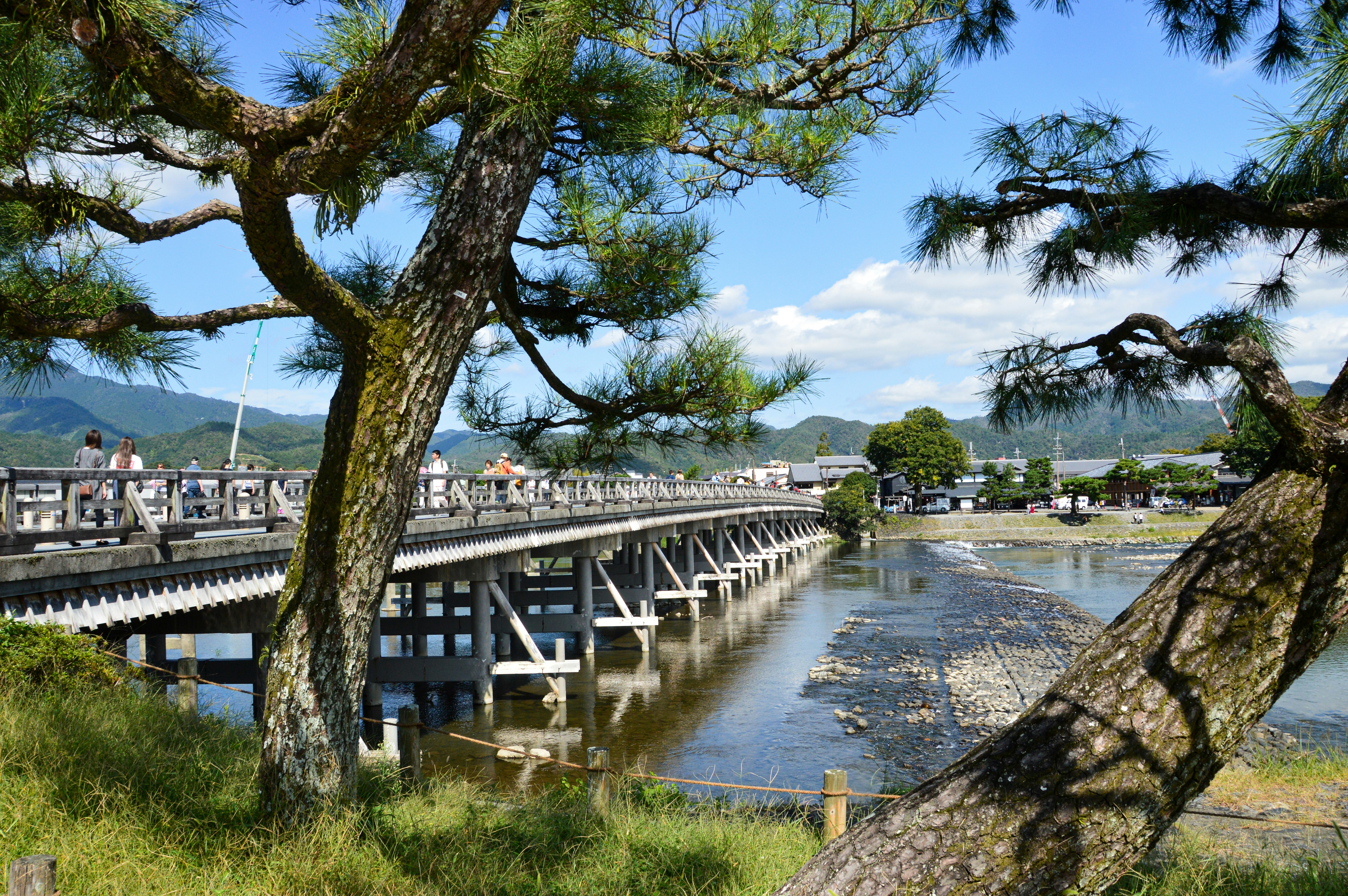 A wooden bridge spans a tranquil river, framed by lush trees and distant hills, showcasing a blend of natural beauty and architectural craftsmanship.