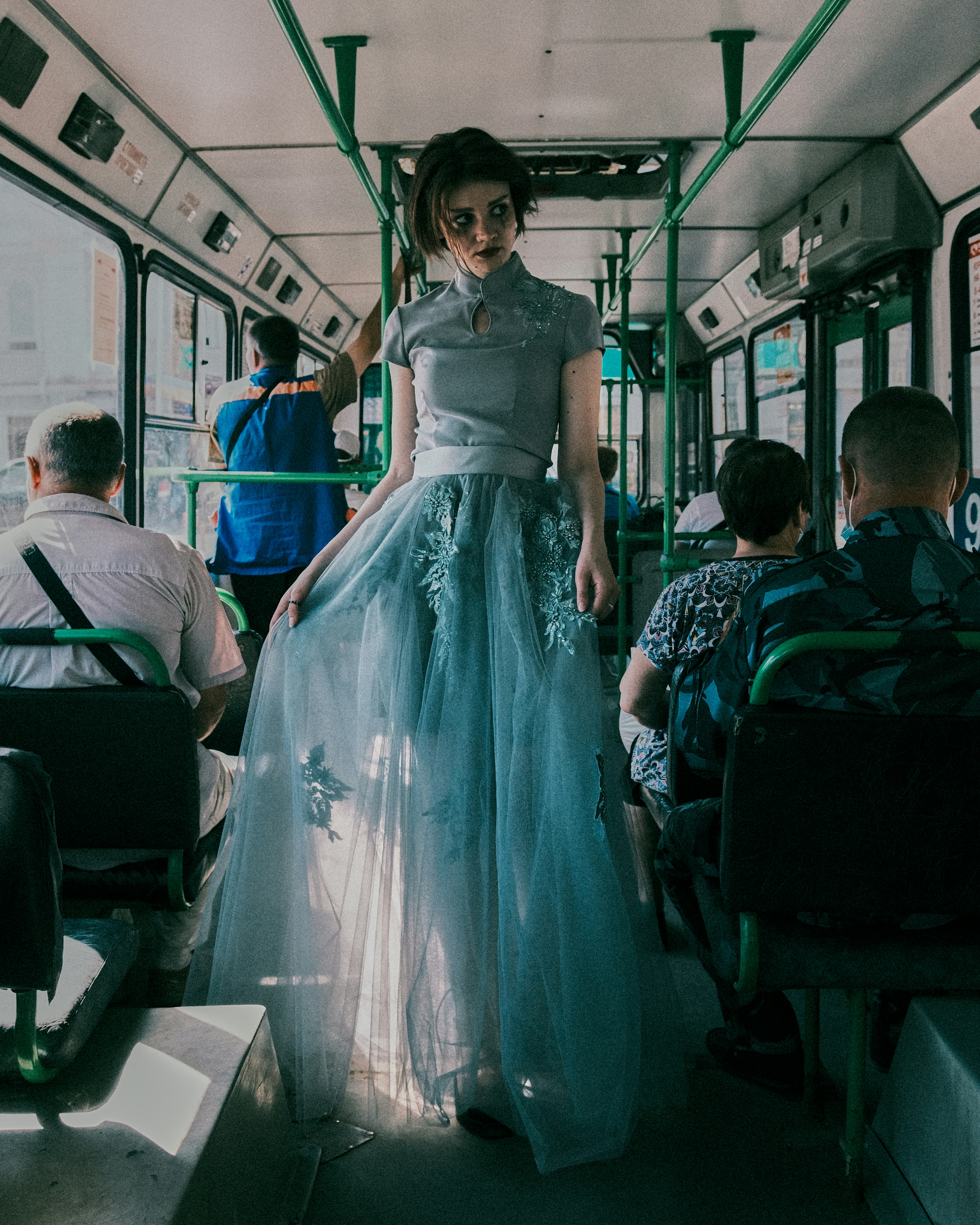 A model in a flowing blue gown stands poised in a crowded bus, contrasting the everyday environment with her high-fashion attire.