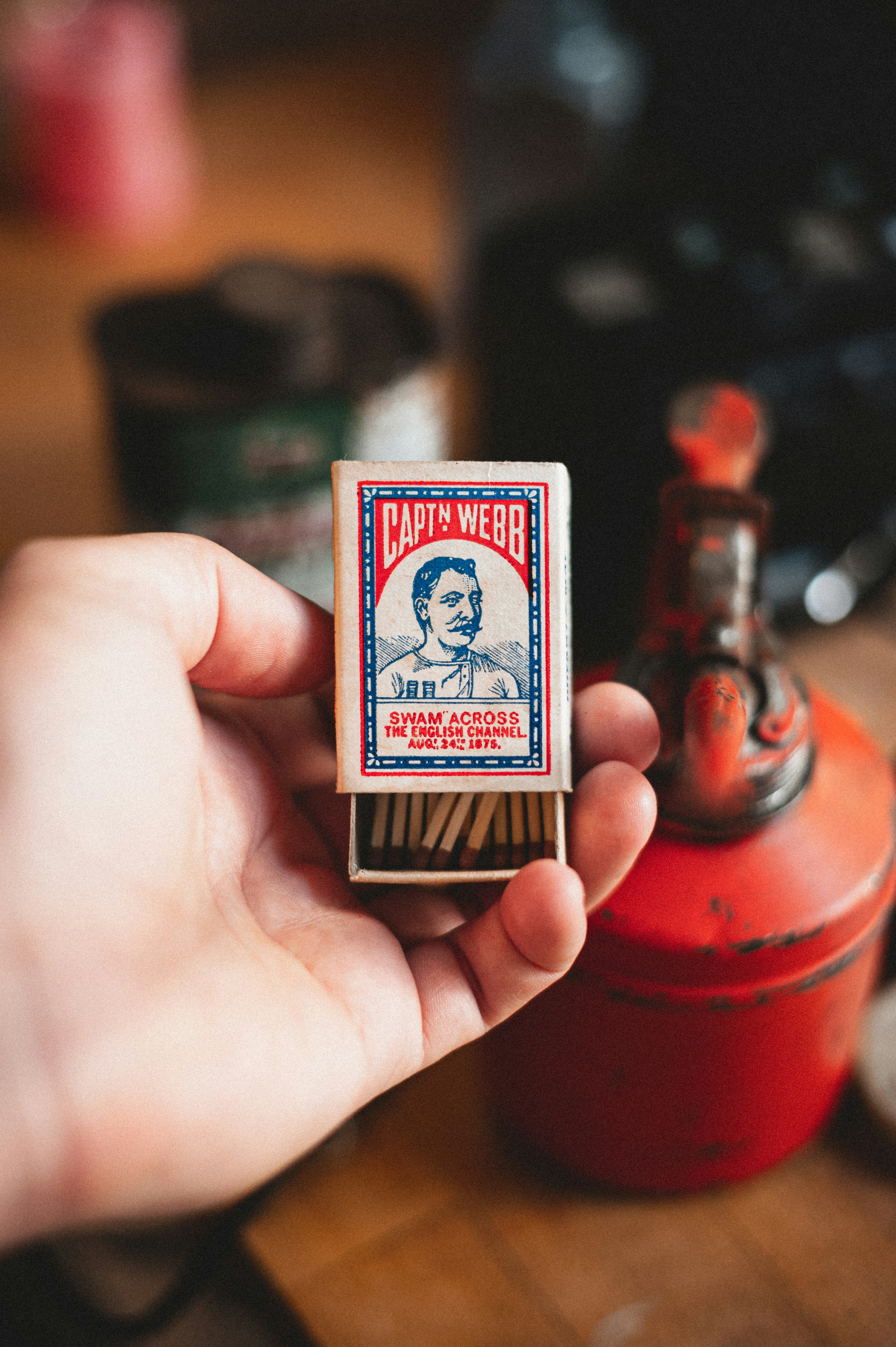 Vintage matchbox featuring Captain Webb, the first person to swim across the English Channel, held in a hand. The background includes a rustic red lantern and various containers.