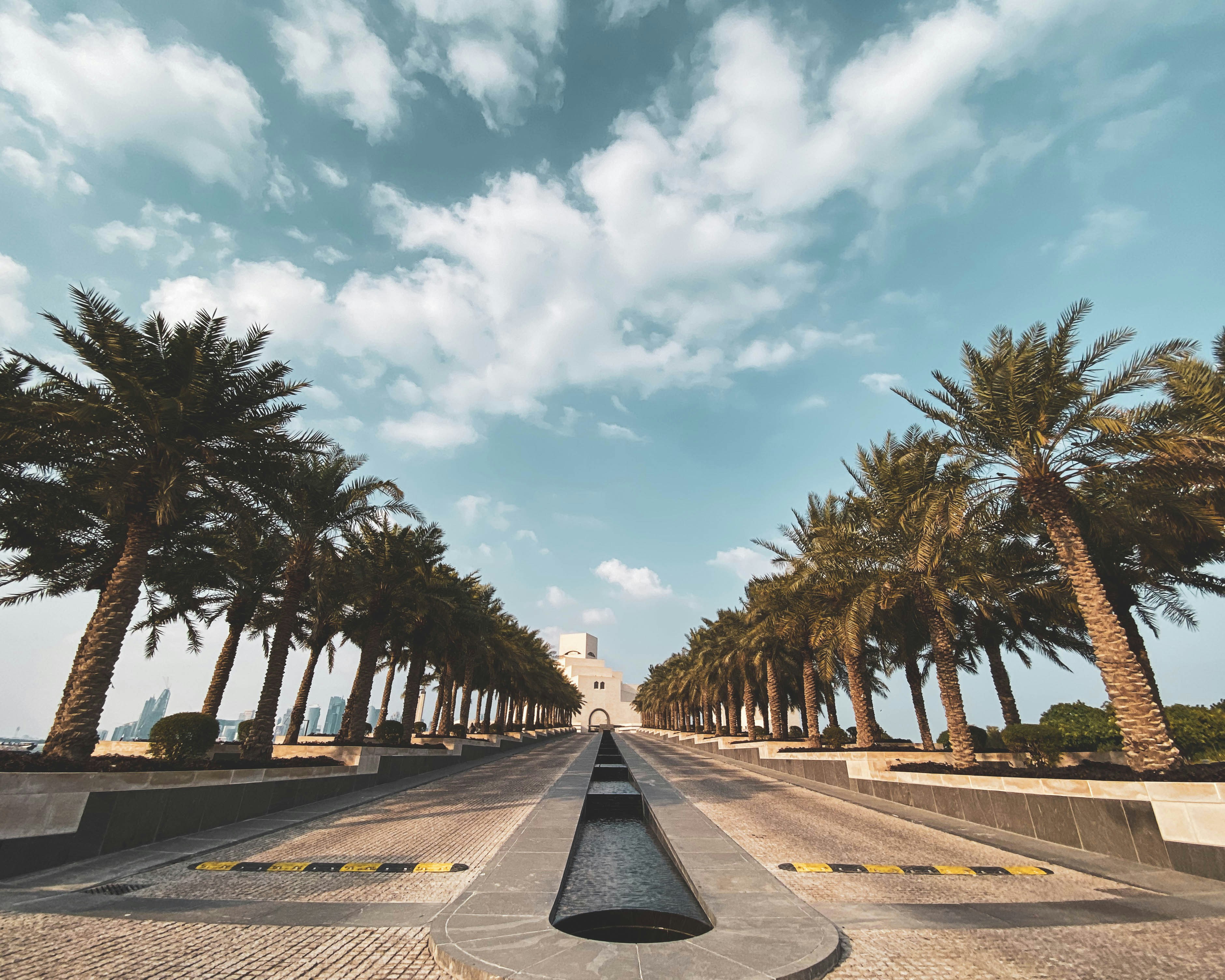 Symmetrical avenue of palm trees leading to a distant monument under a vibrant blue sky with scattered clouds.
