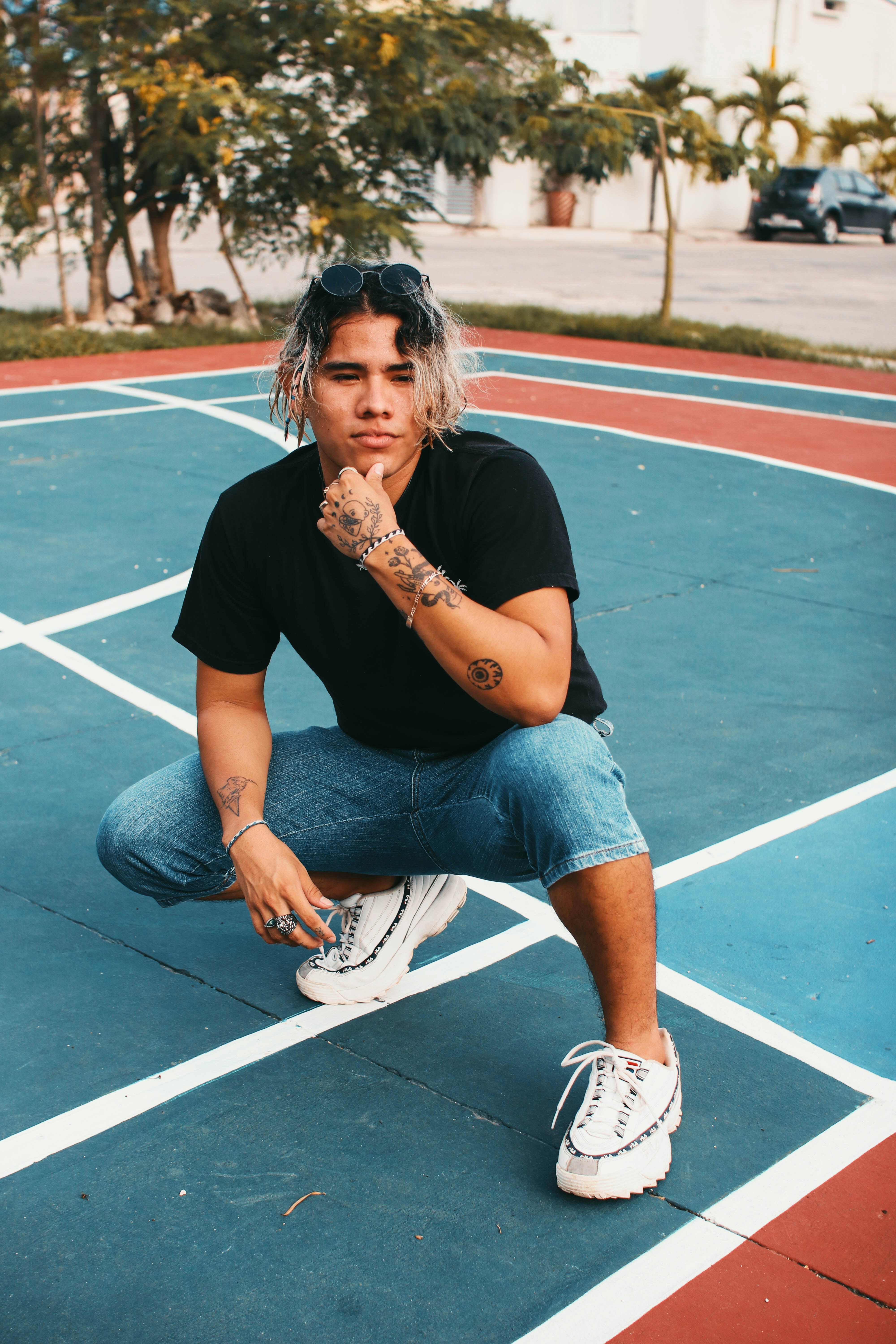 Portrait photograph of a tattooed man crouching on a colorful basketball court. He wears a black t-shirt, denim shorts, white sneakers, and sunglasses perched on his head.