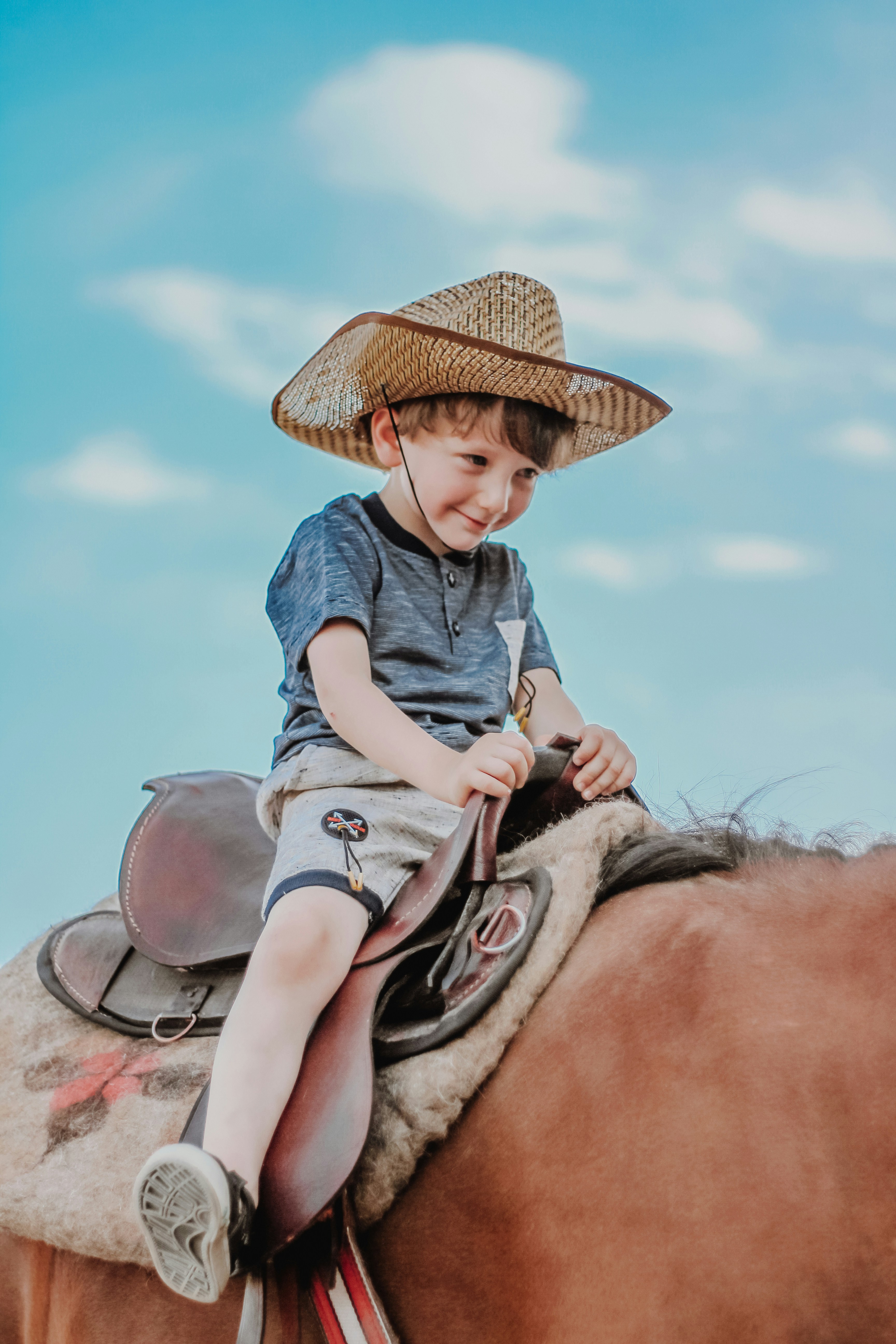 Young boy in a straw hat rides a horse, radiating happiness against a bright blue sky.