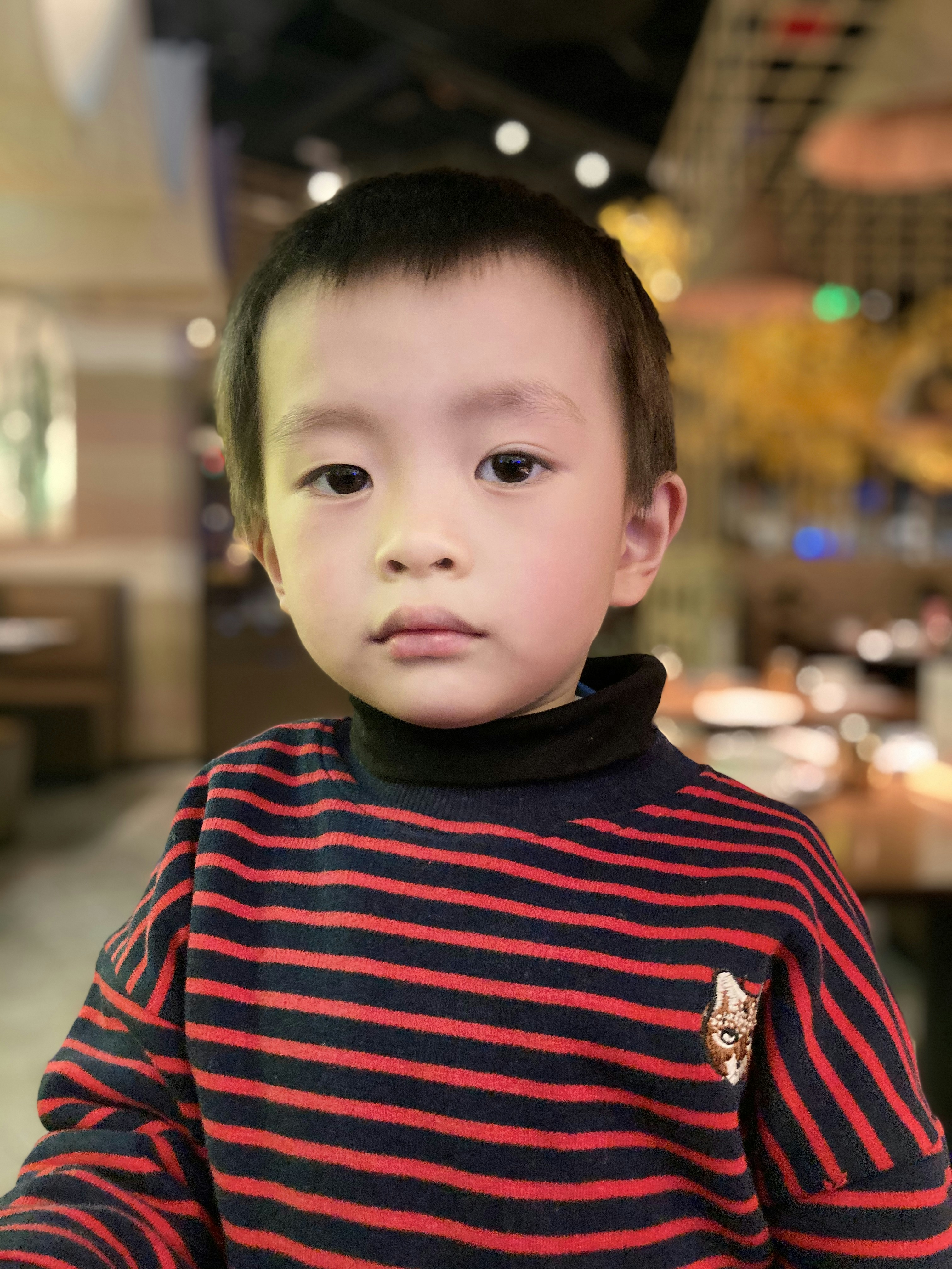 Young child with a serious expression, wearing a striped sweater with a small animal emblem, set against a softly blurred restaurant background.