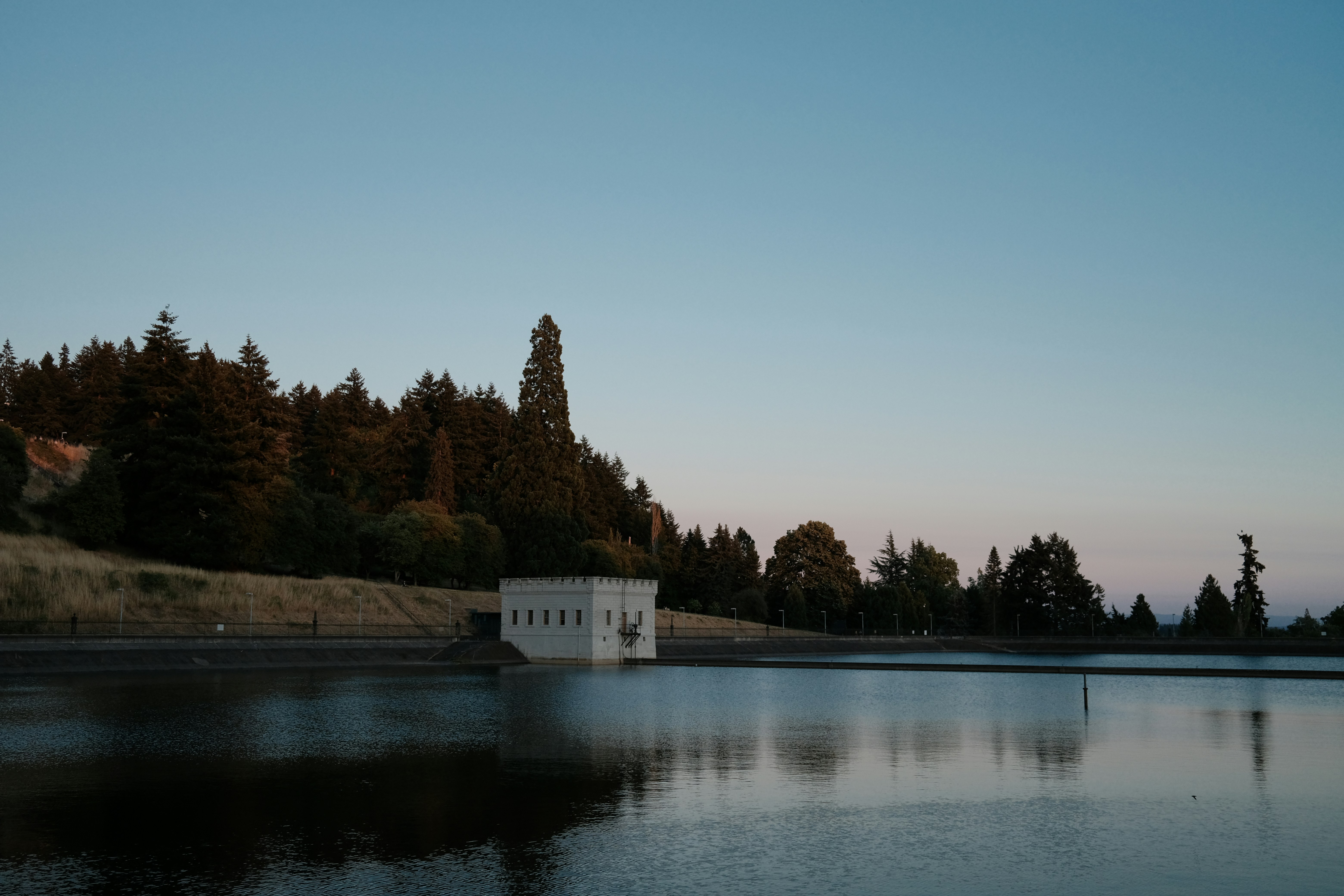 Tranquil reservoir with a small building, surrounded by trees at dusk.