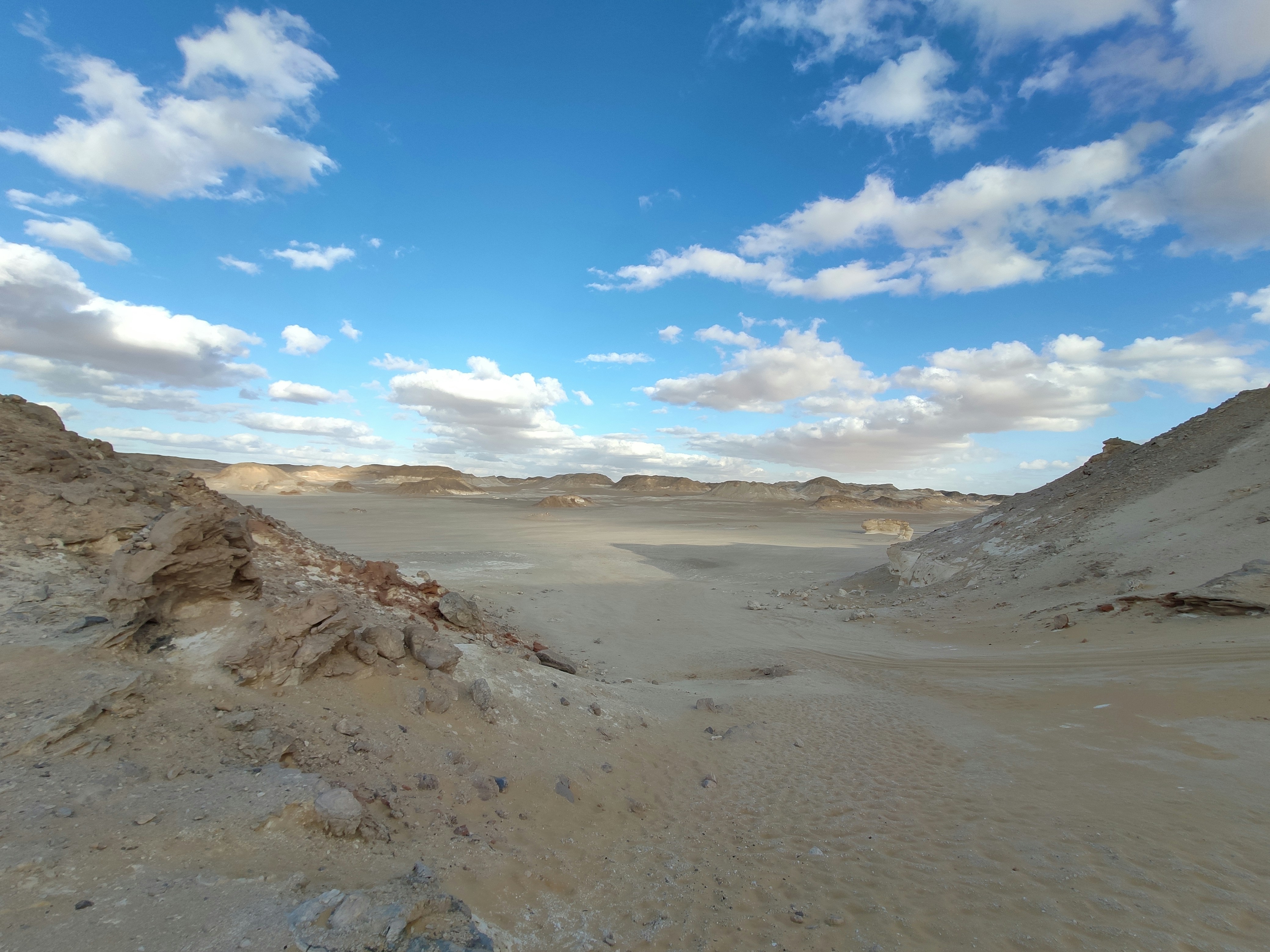 Expansive sandy landscape with rocky formations under a blue sky dotted with clouds.