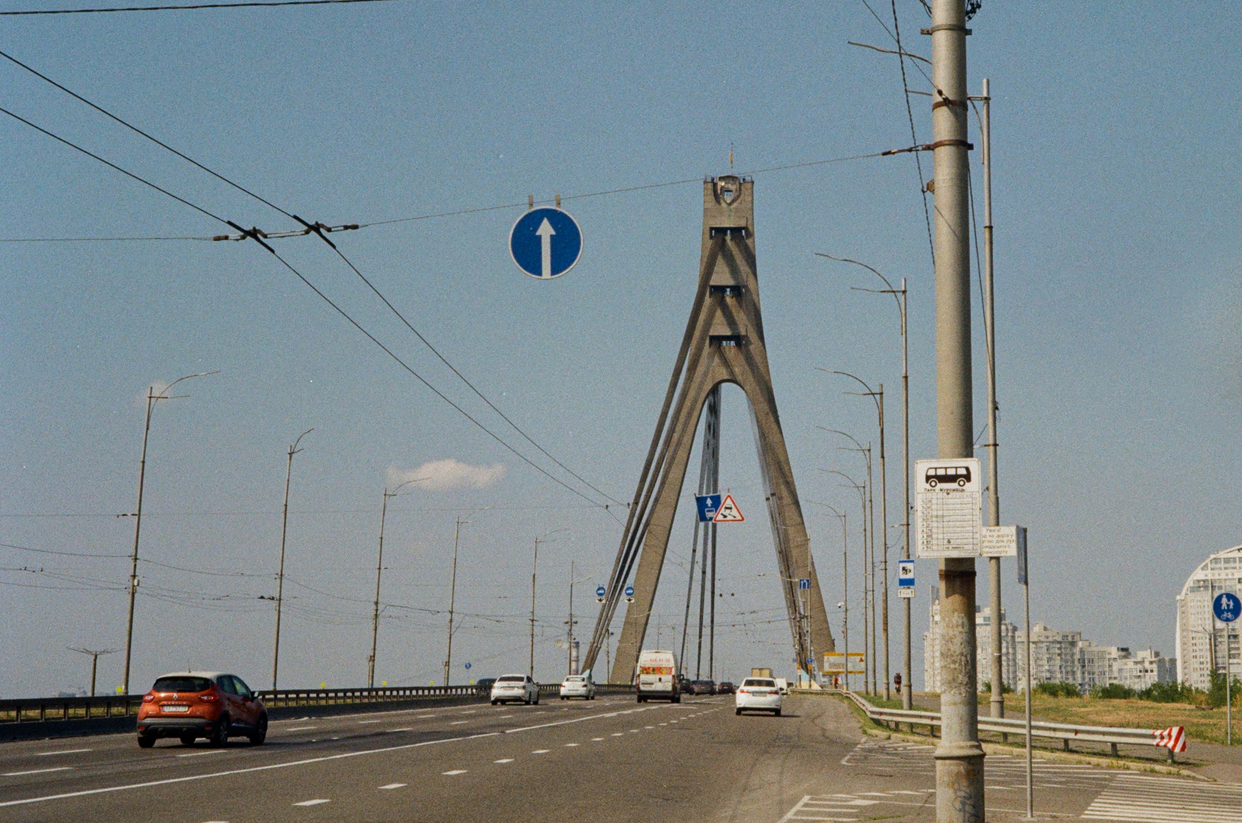 A modern bridge structure rising above a busy road, with traffic signs and cityscape in the background.
