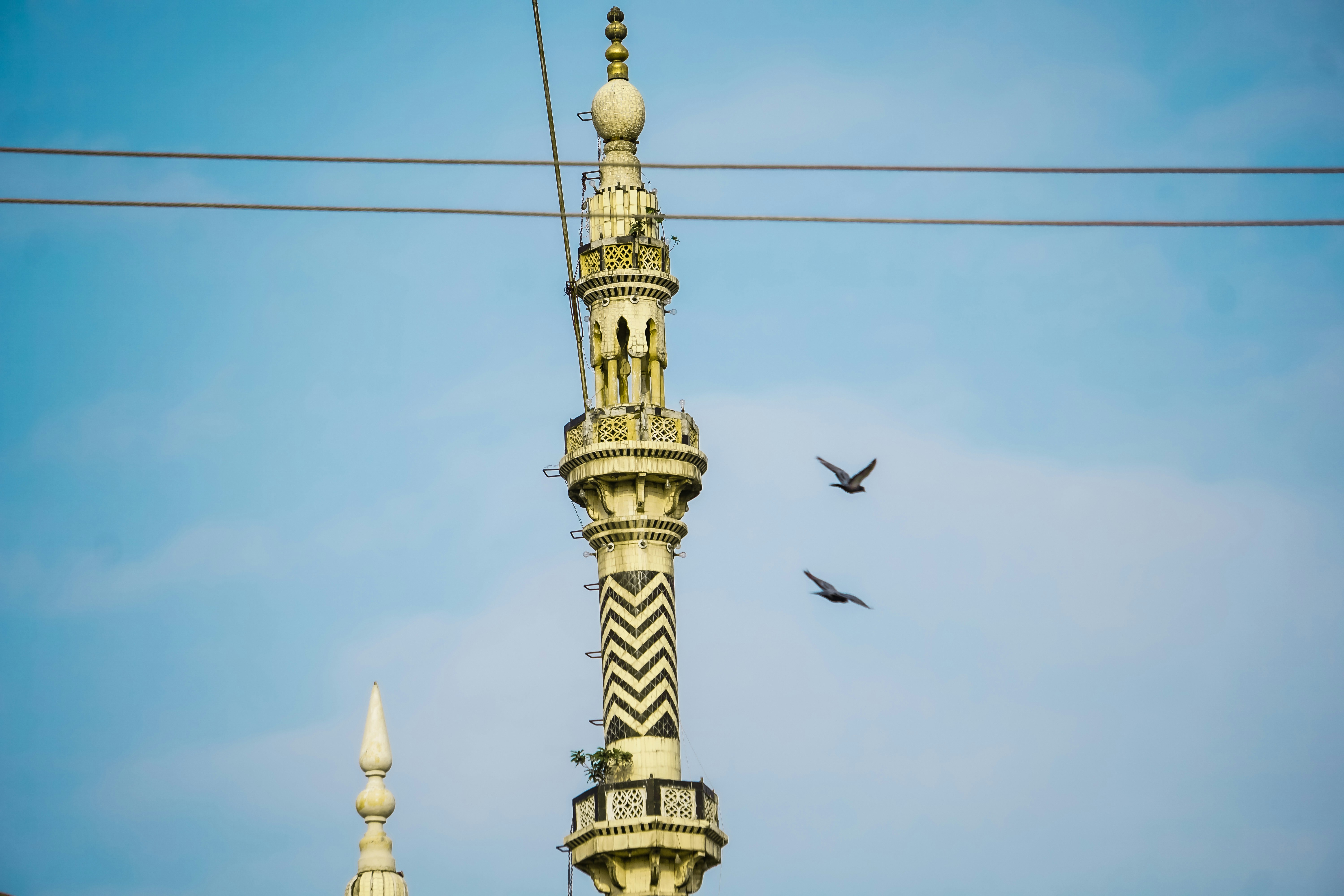 Ornate minaret with intricate patterns set against a clear blue sky as birds fly nearby.