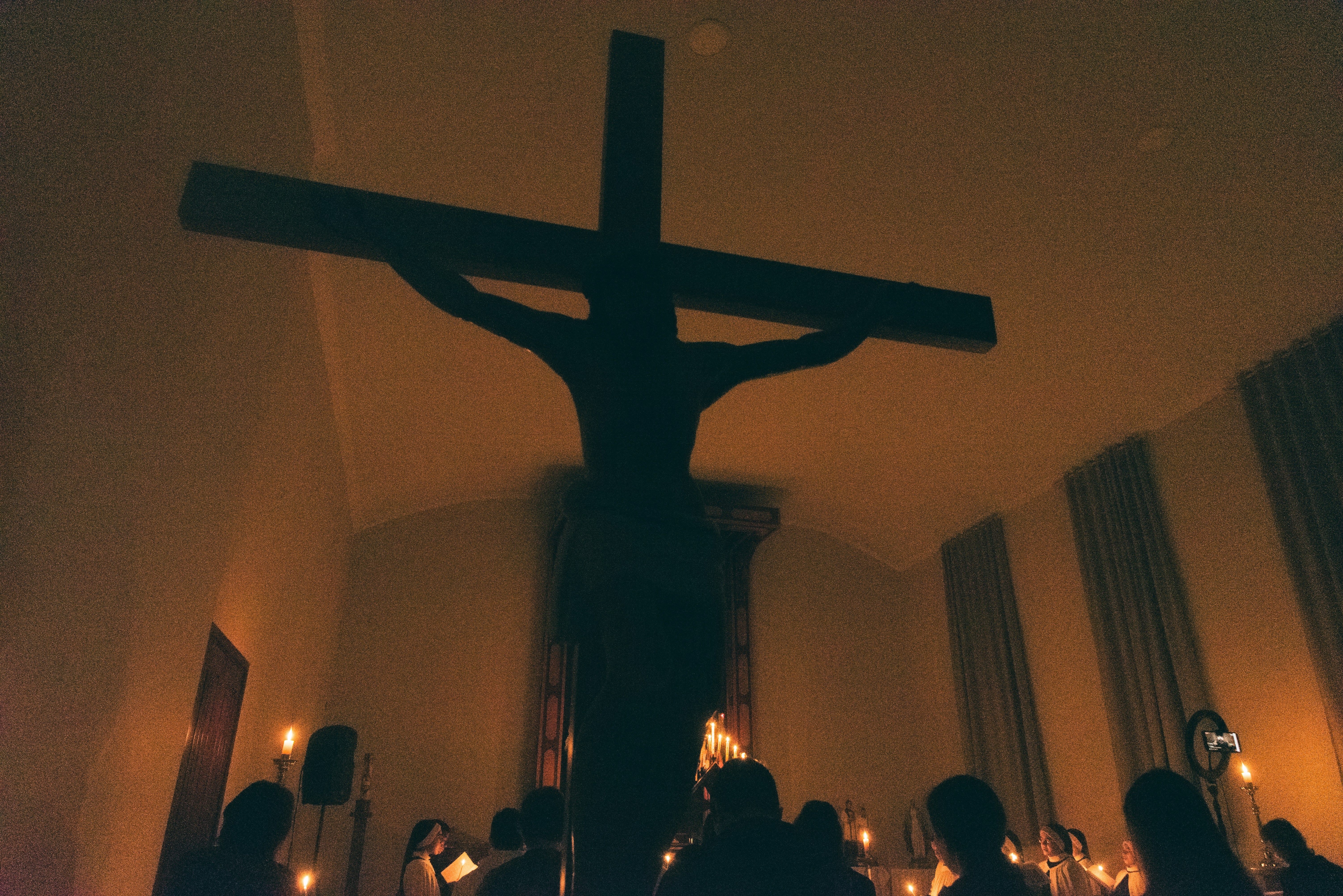Silhouette of a large crucifix surrounded by candlelit worshippers in a dimly lit room.