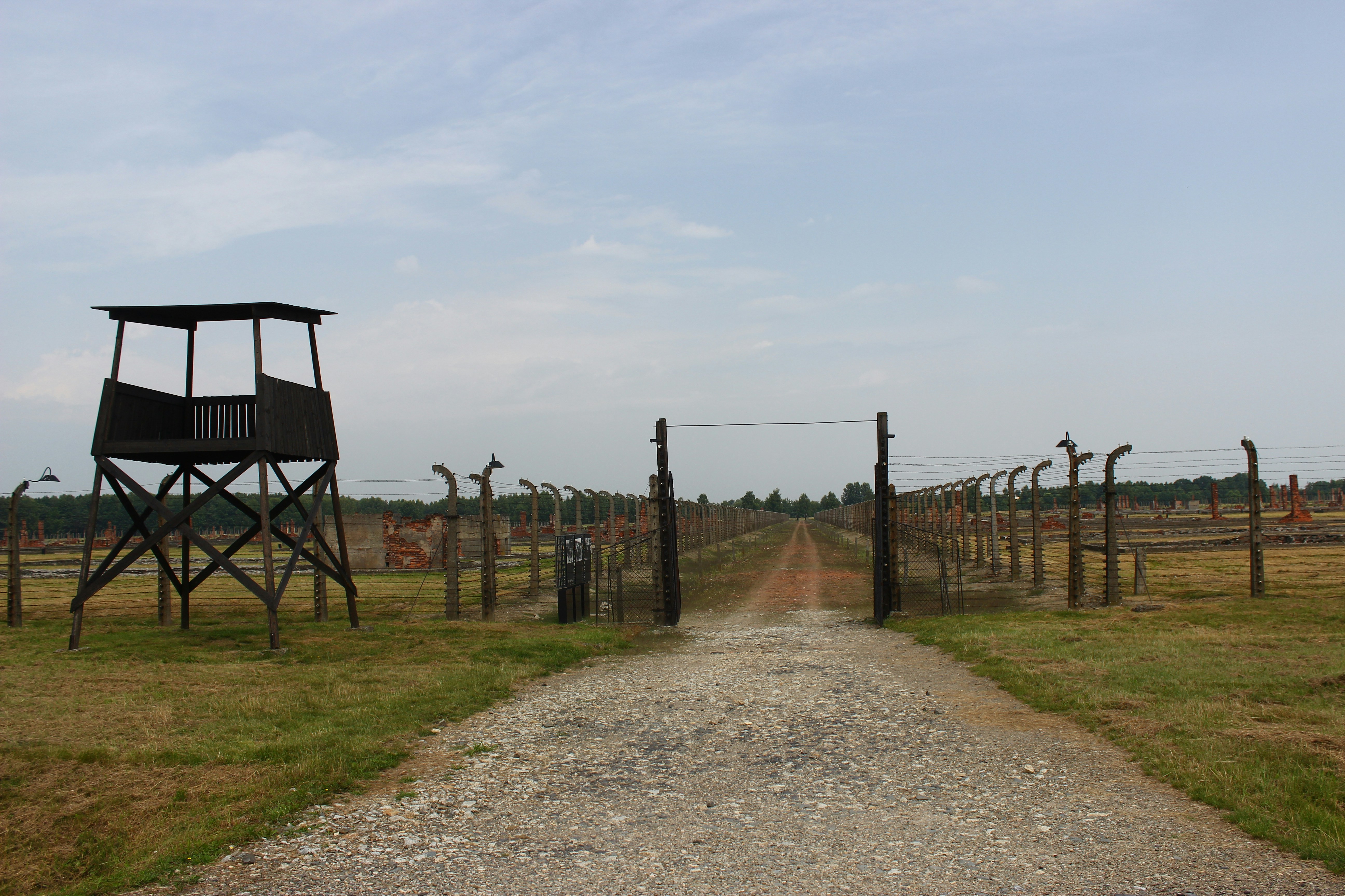 Gravel path leading through a metal fence with a watchtower on a grassy field under a cloudy sky.