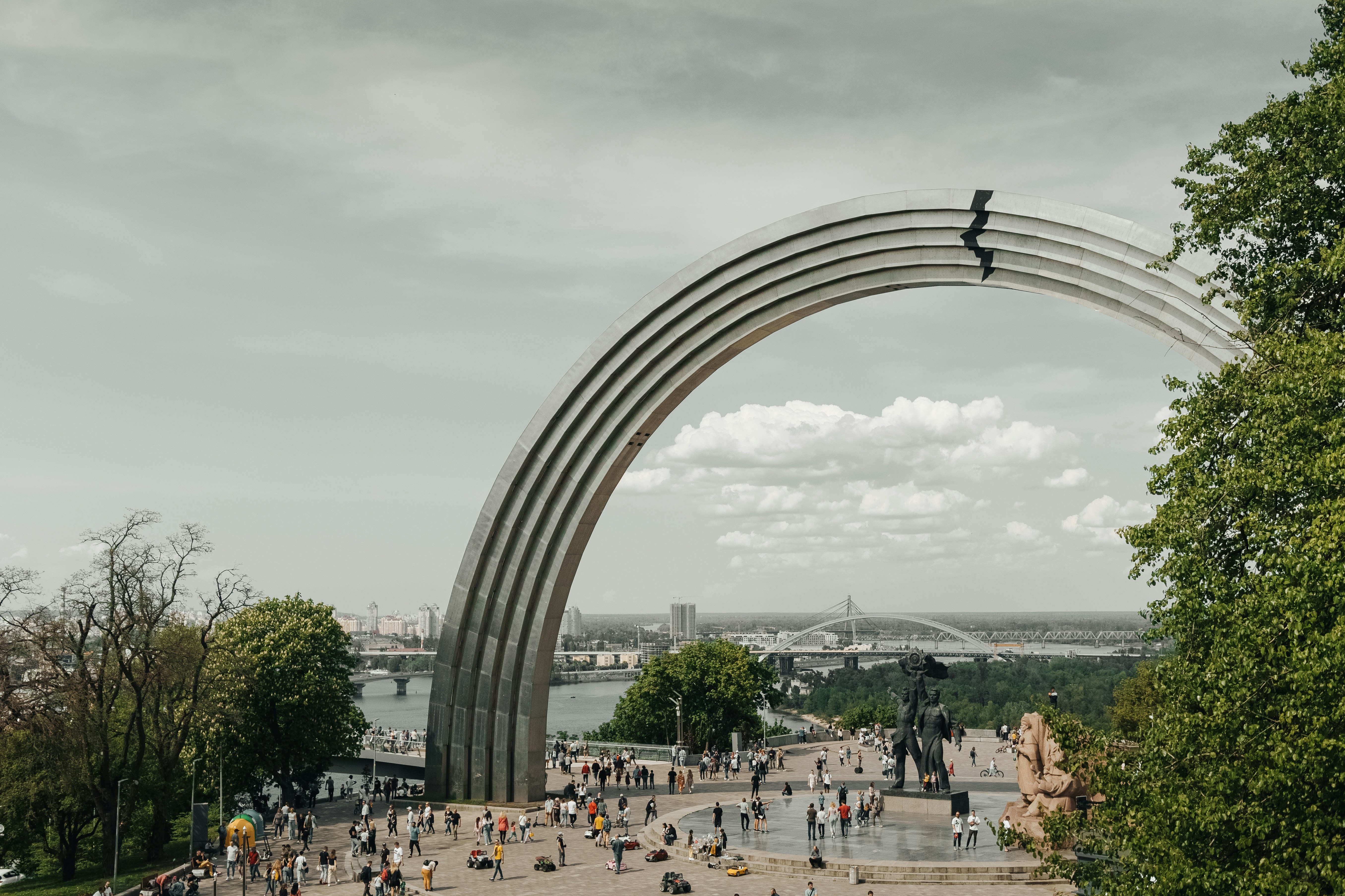 Public plaza beneath a multi-ring metal arch with crowds and a distant skyline. The composition highlights the arch as a landmark within a busy urban scene.