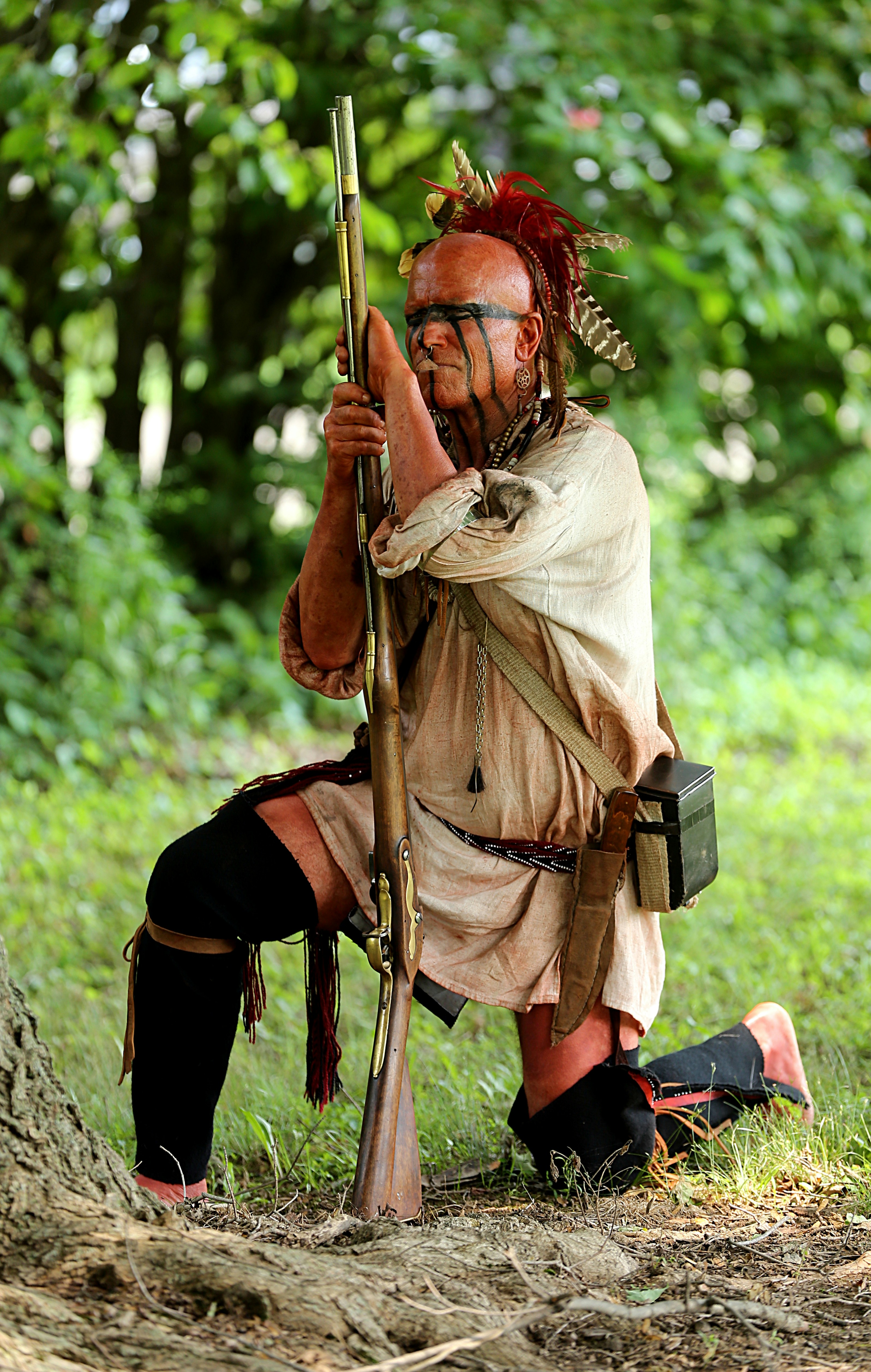 A figure in traditional attire kneels with a vintage rifle, embodying a moment of focus amidst a lush green backdrop. The scene captures a connection to heritage and nature.