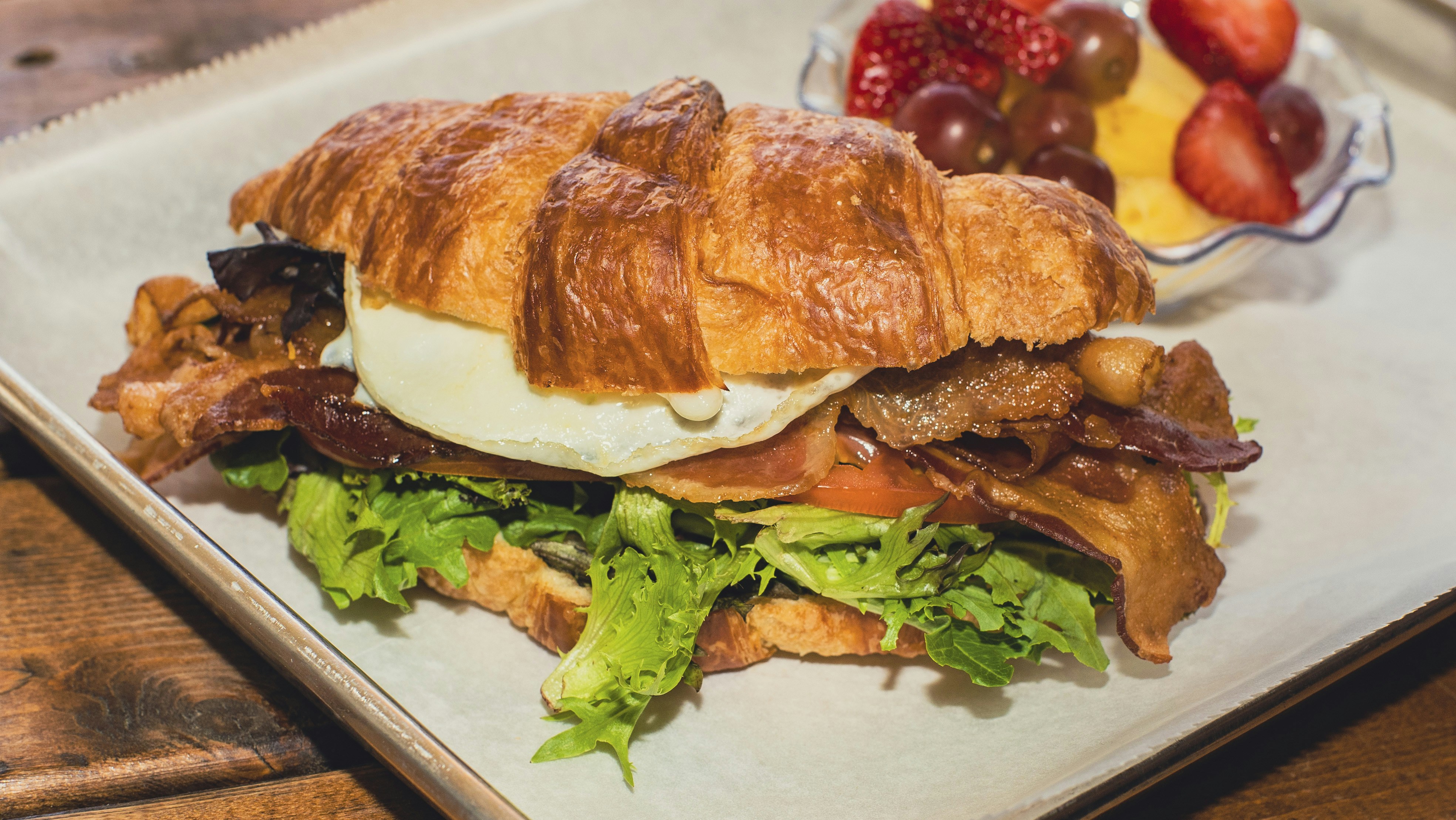 Croissant sandwich filled with crispy bacon, lettuce, and a fried egg, accompanied by a bowl of mixed fruit.