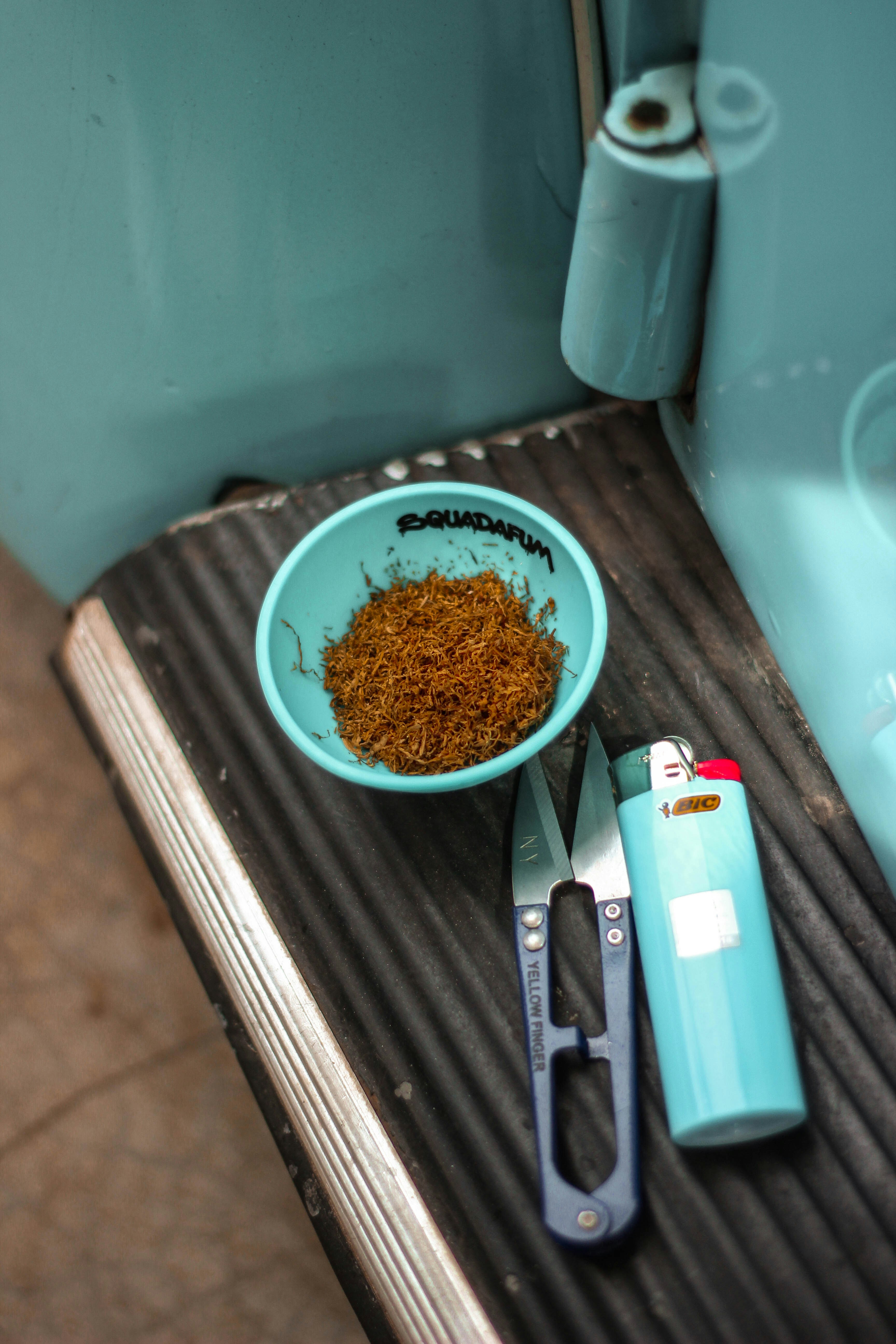 Teal Bowl of Shredded Tobacco Amid a Folding Knife and Cyan Lighter in a Pickup Bed