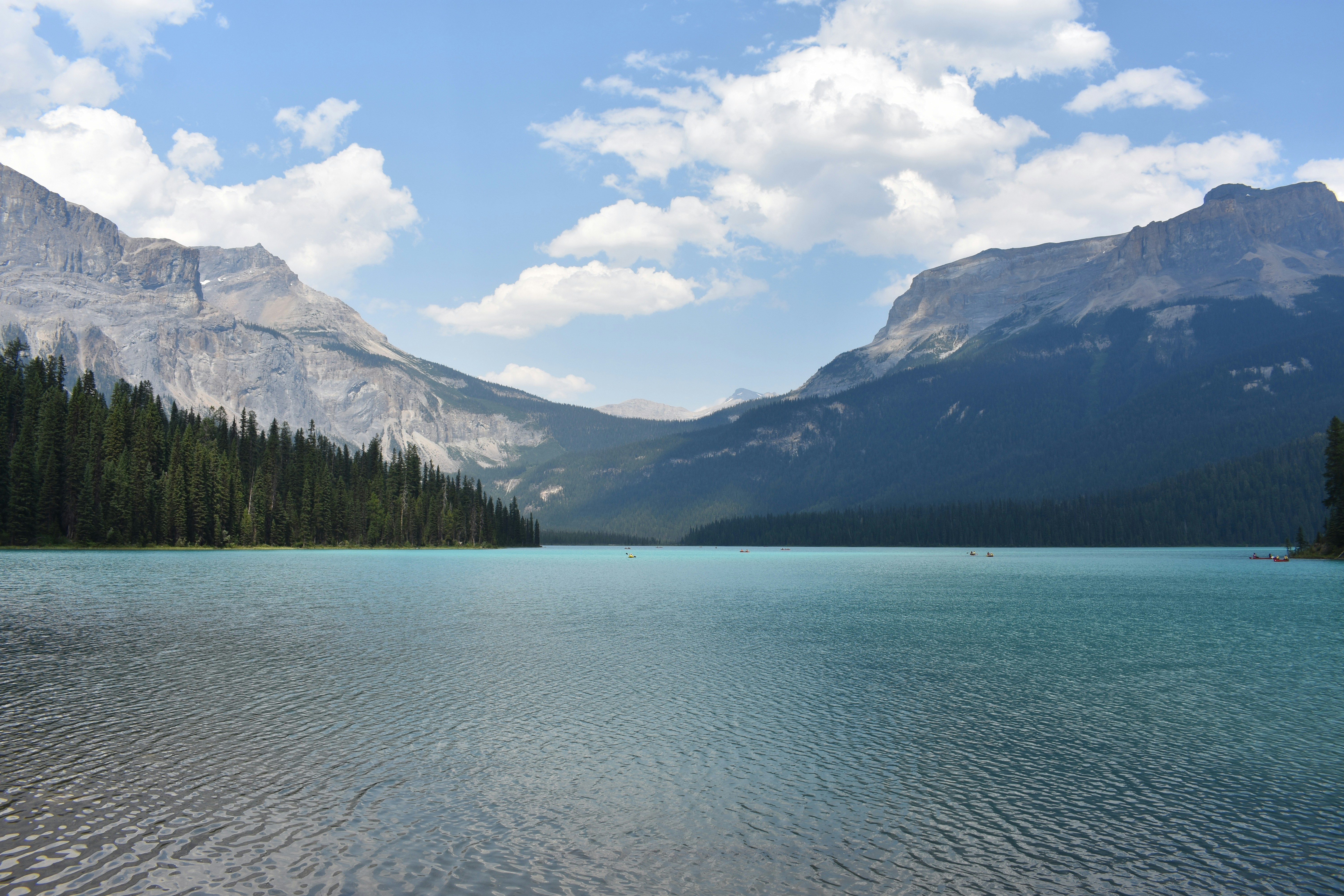 Clear blue lake bordered by dense forest with towering mountains under a partly cloudy sky.