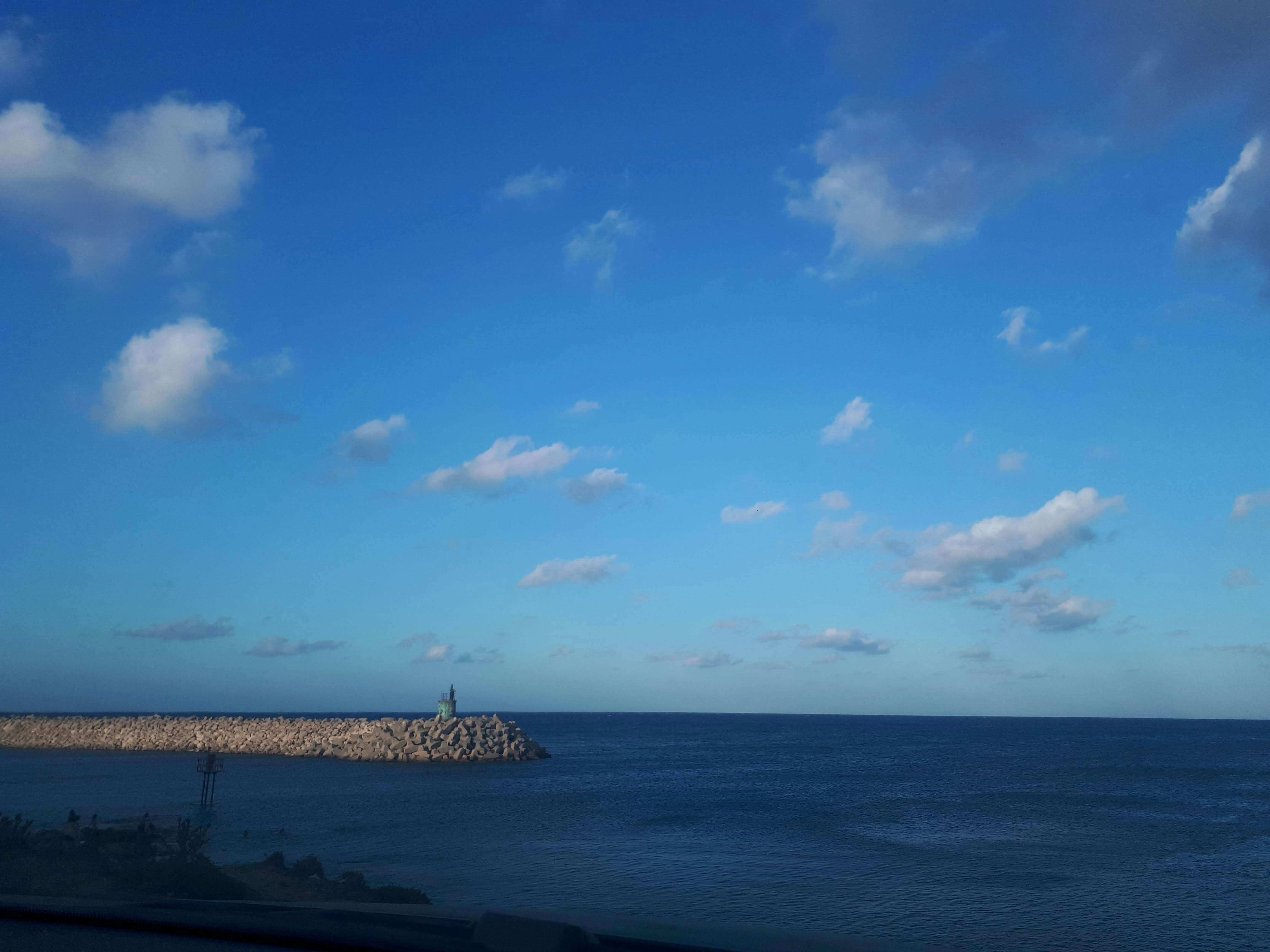 Calm seascape with a low breakwater extending toward the horizon and a distant lighthouse under a crisp blue sky.
