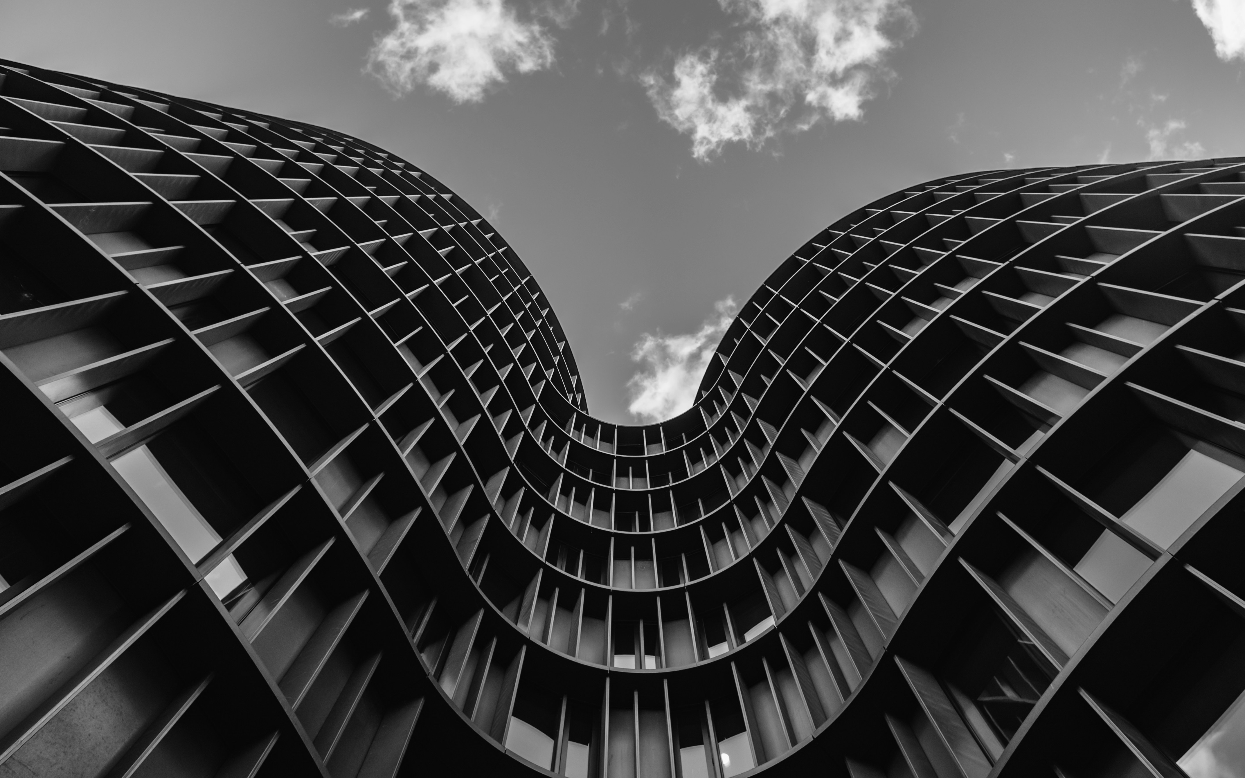 Grayscale photo of a curving, modern concrete building with a dynamic facade under a cloudy sky.