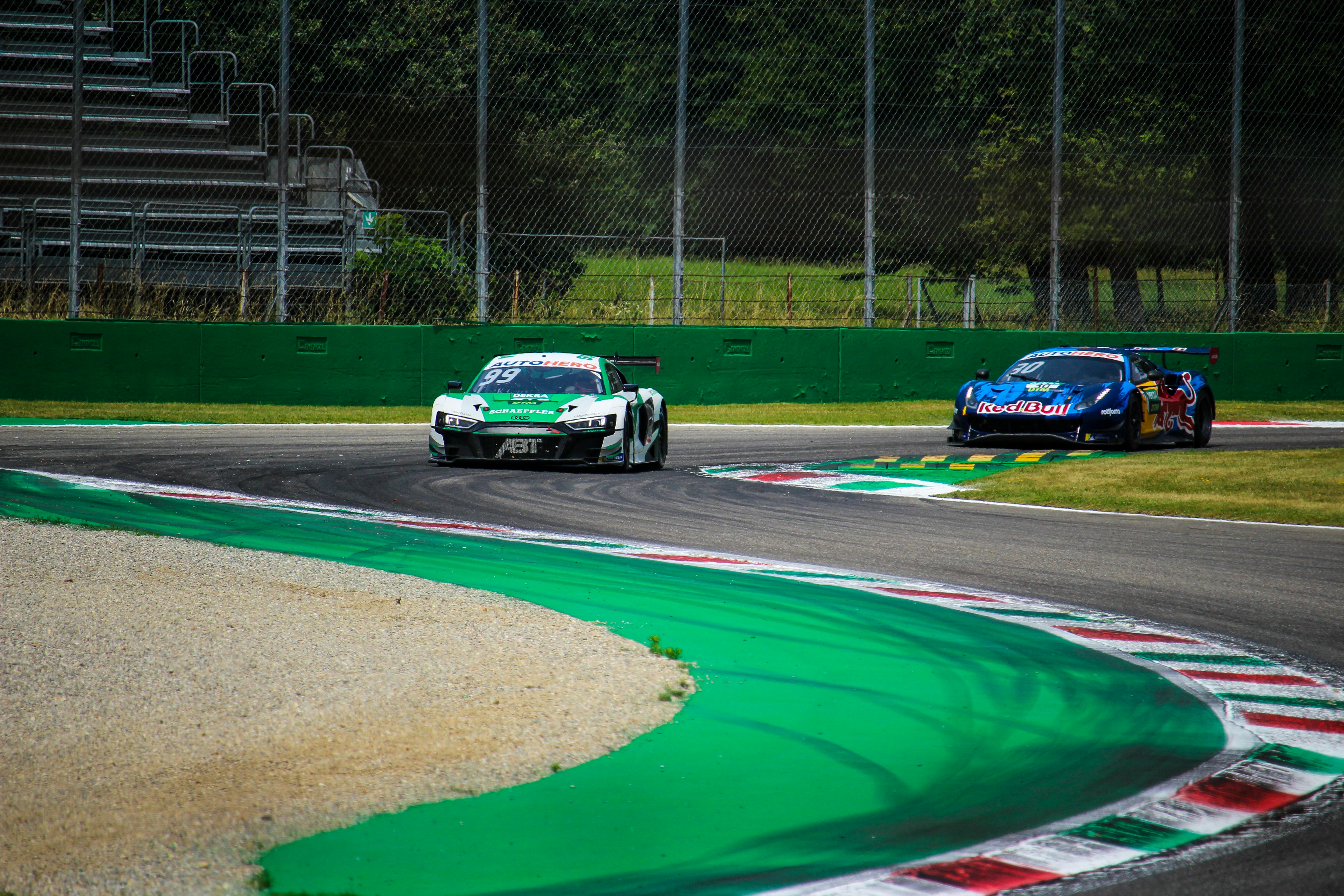 Two sports cars navigate a sharp racetrack curve, one white and one blue, with lush greenery in the background.