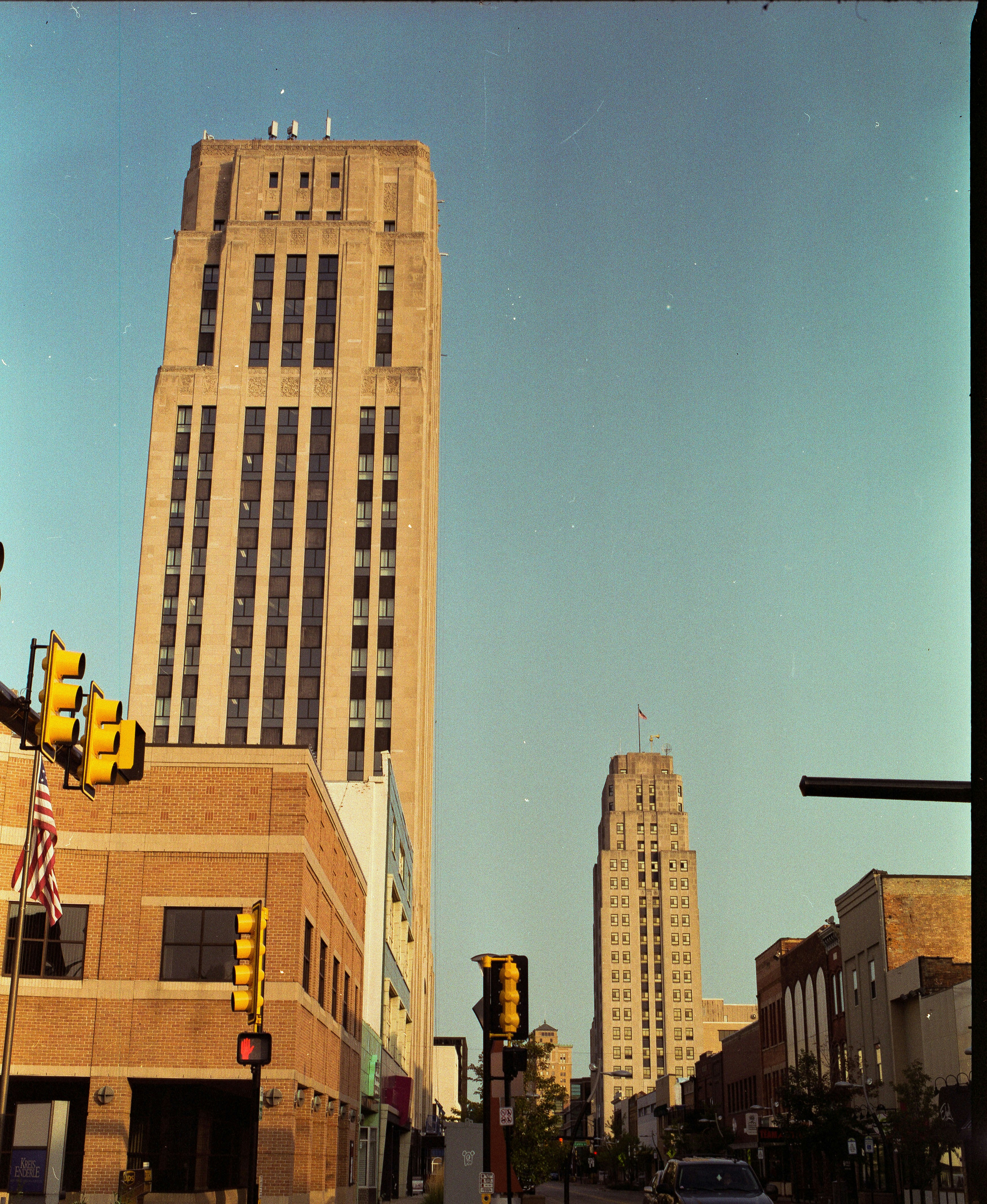 Three prominent buildings rise against a clear blue sky, showcasing a blend of modern and historical architecture. Traffic signals and storefronts add urban context.