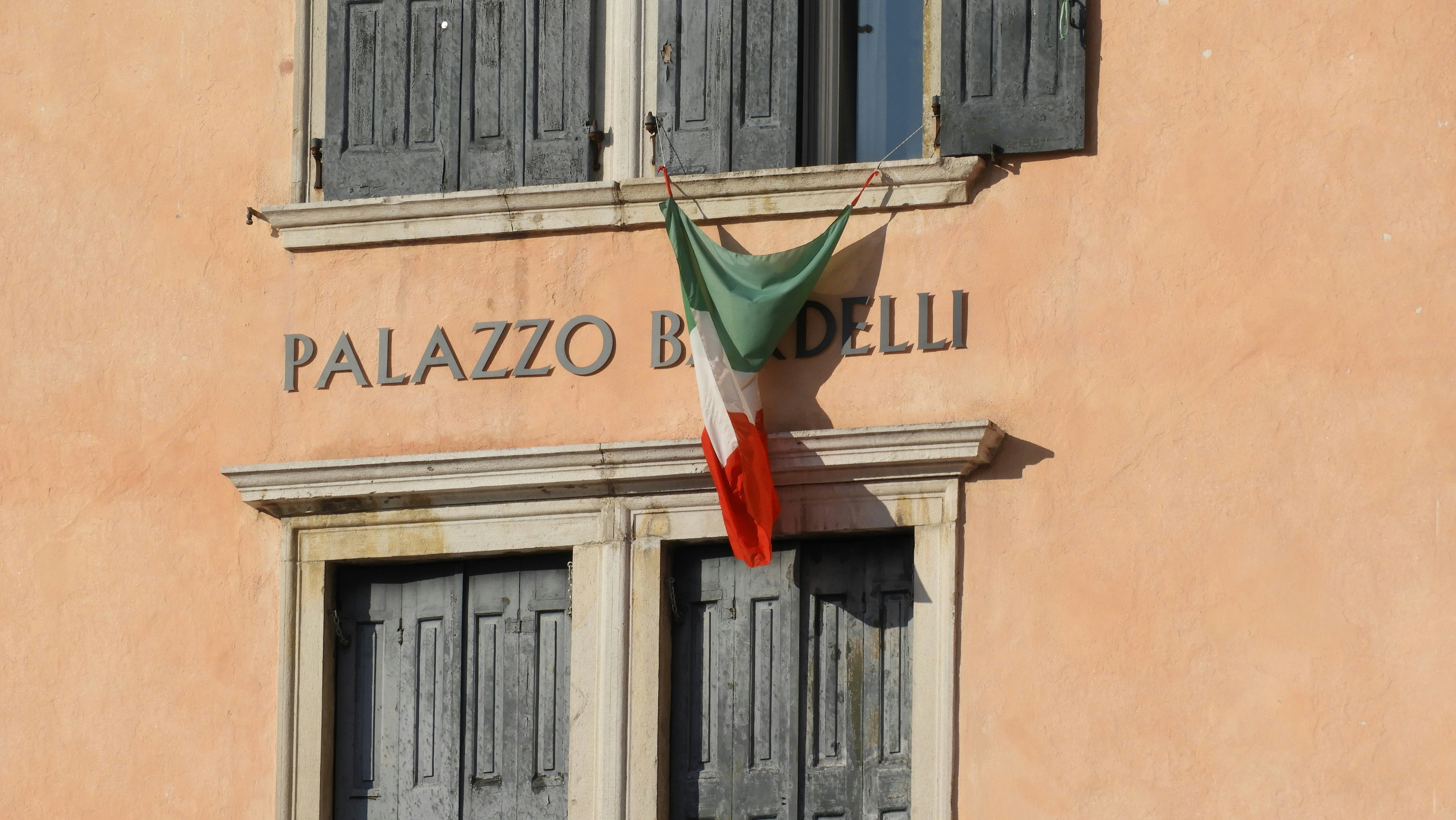 Flag of Italy draped from a window of Palazzo Bradelli, showcasing the building's historic charm against a warm peach facade.
