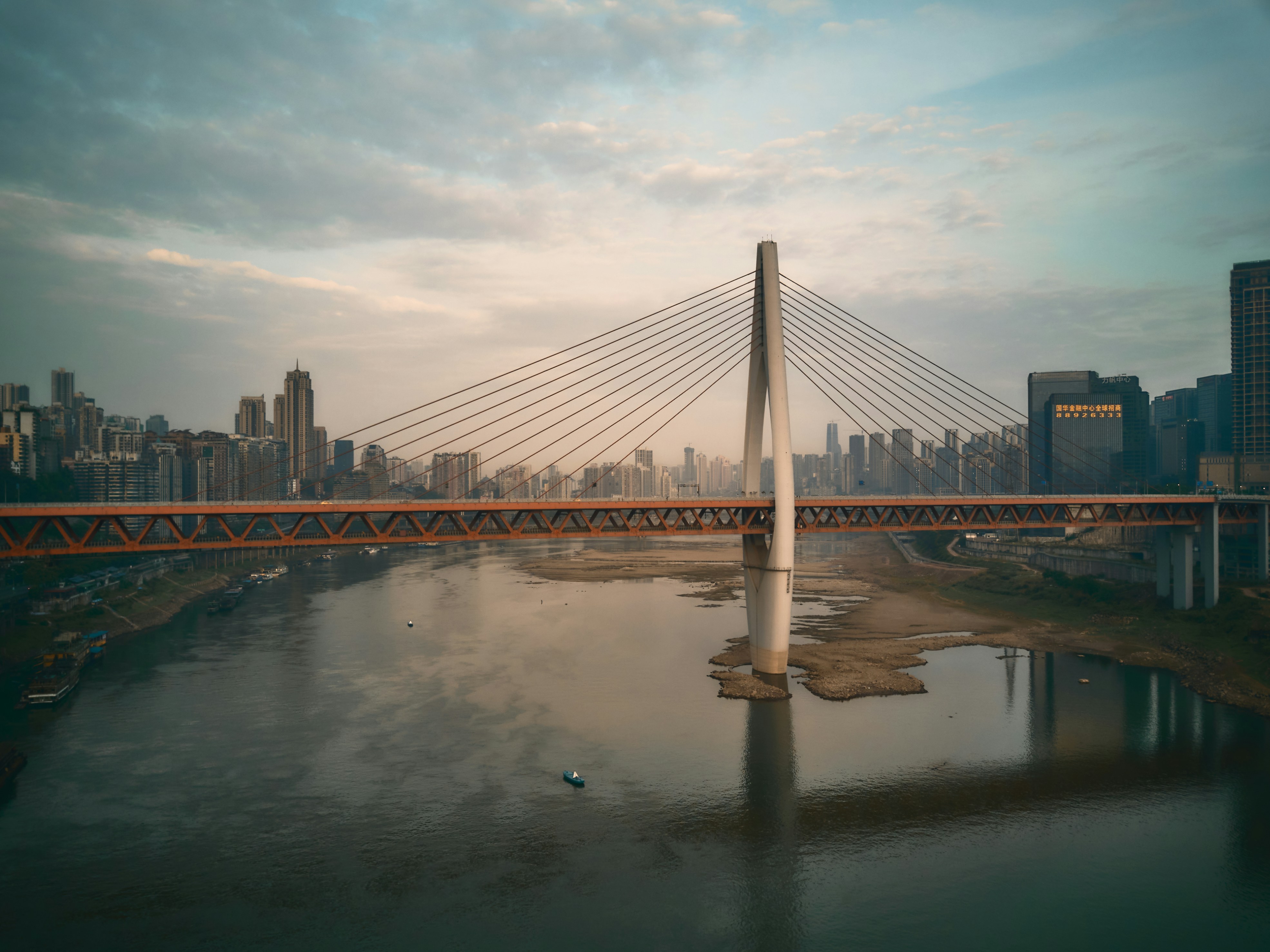 Cable-stayed bridge stretches across a calm river with a city skyline under a cloudy sky.
