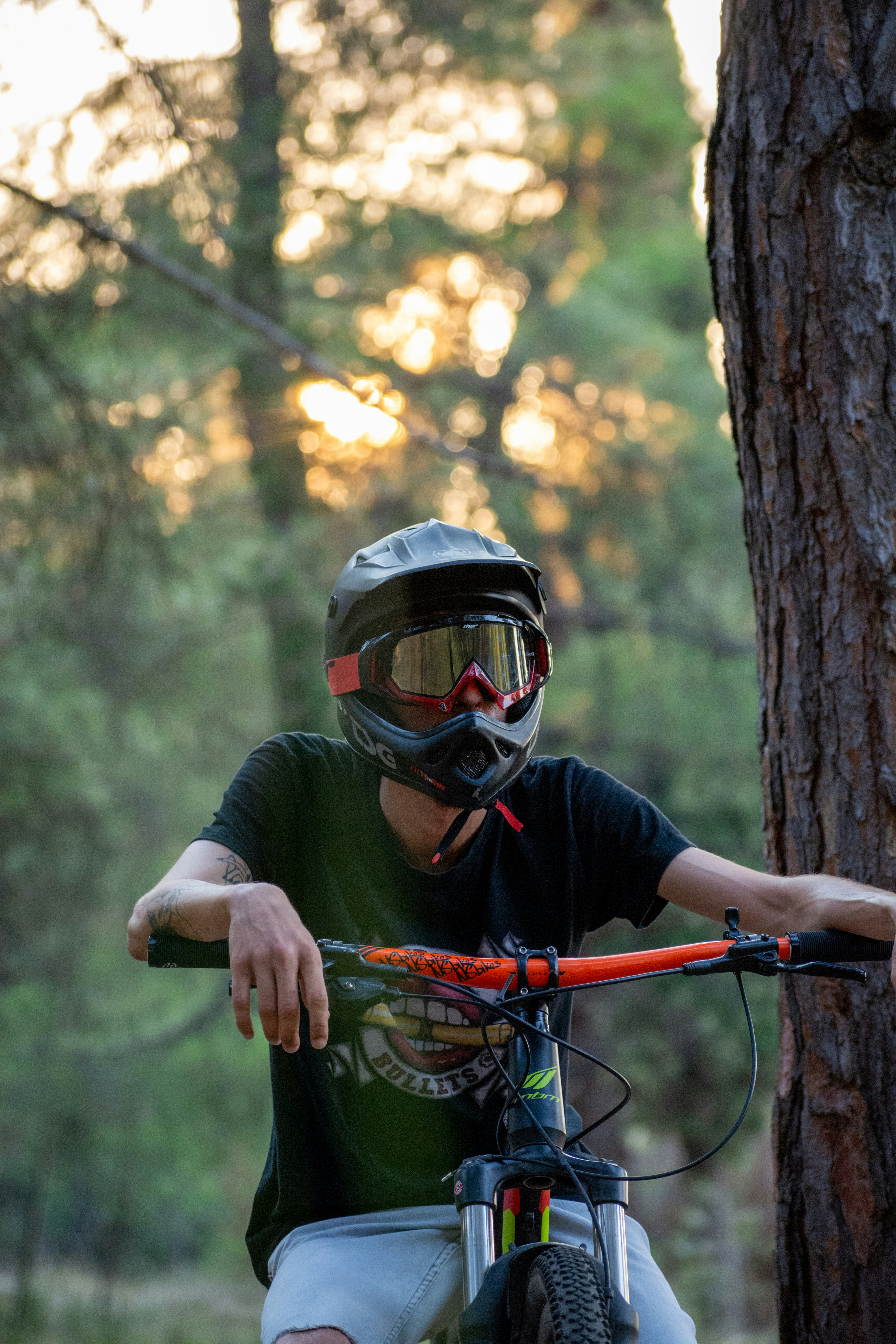Mountain biker in protective gear rests against a tree, with a softly blurred sunset backdrop illuminating the scene.