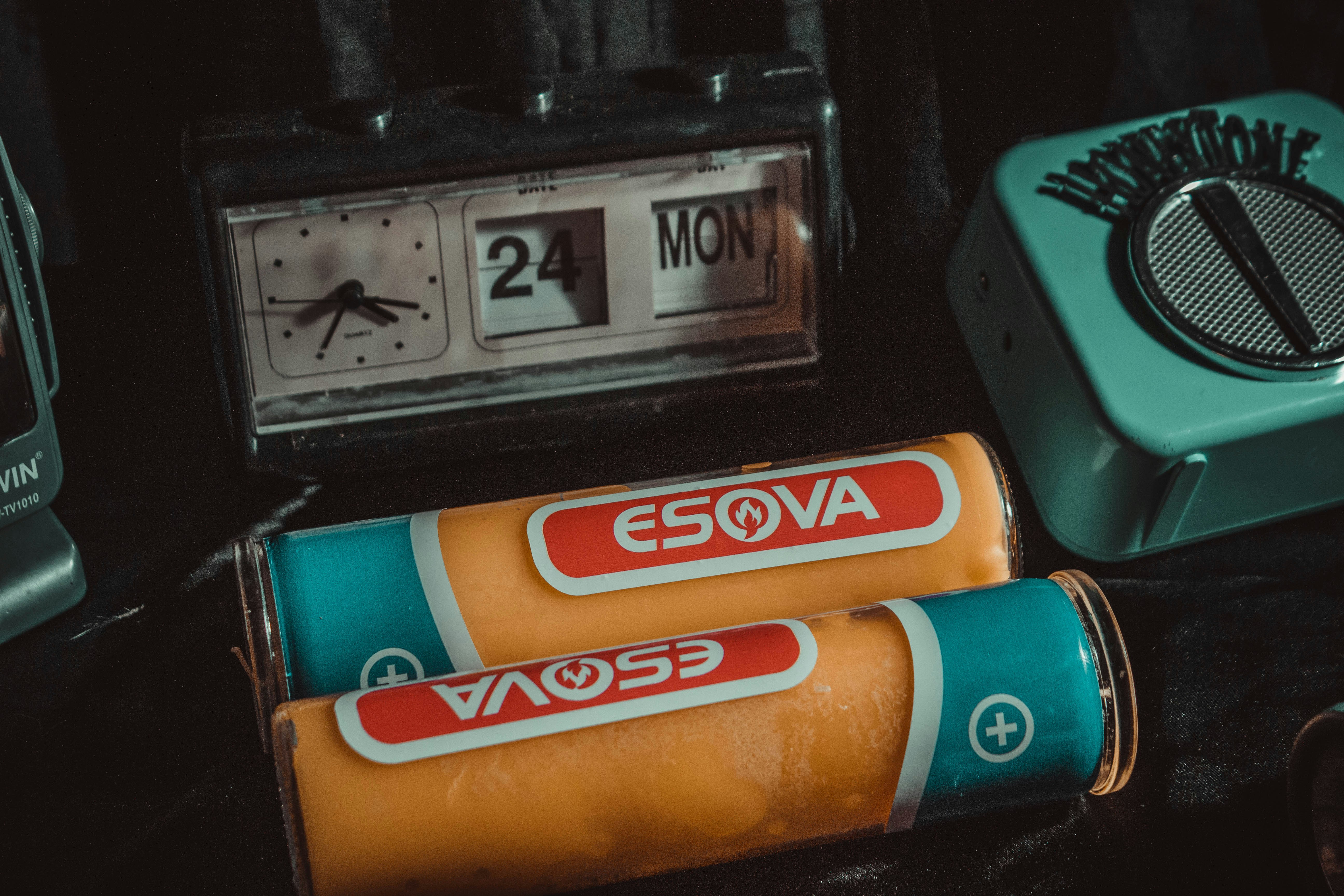 Two vintage batteries with the label 'ESOVA' positioned alongside a classic clock and a teal speaker, evoking nostalgia for simpler times.