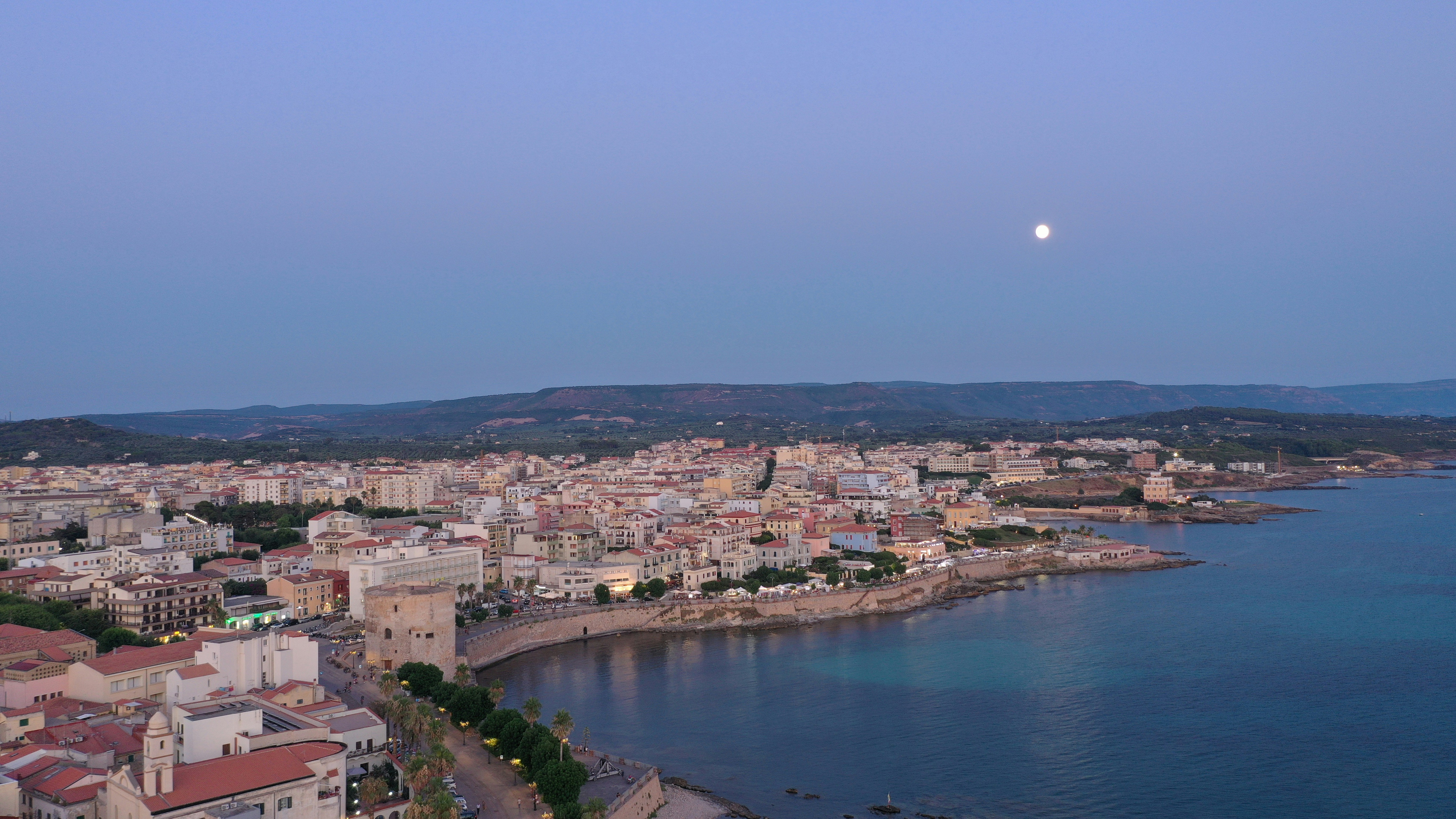 A tranquil coastal town bathed in soft twilight hues, with the moon casting a gentle glow over the water and buildings. The scene captures the peaceful blend of urban life and nature.