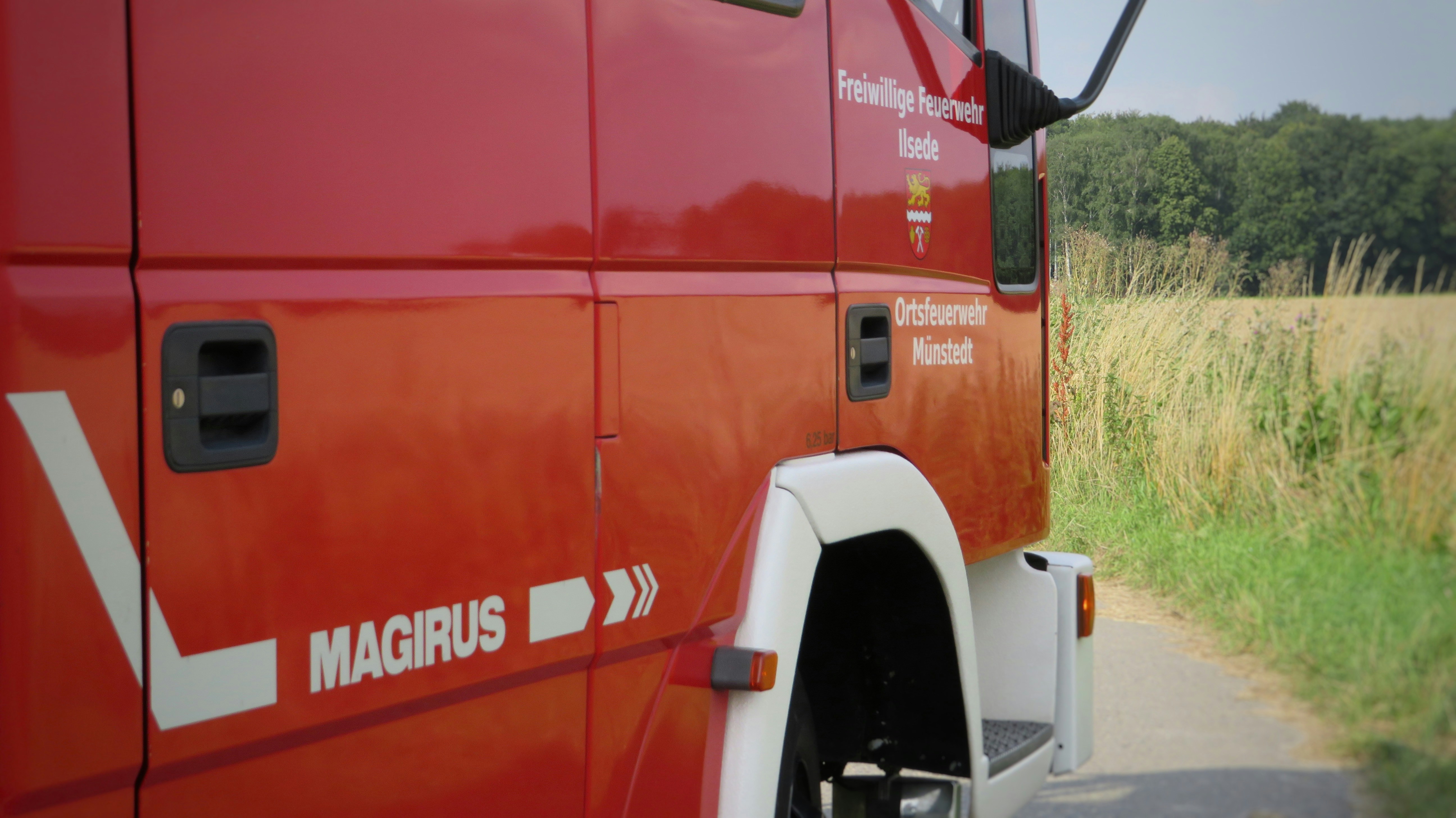 A red fire truck parked beside a rural road, showcasing its distinctive branding and design elements. The surrounding landscape features tall grass and trees in the background.