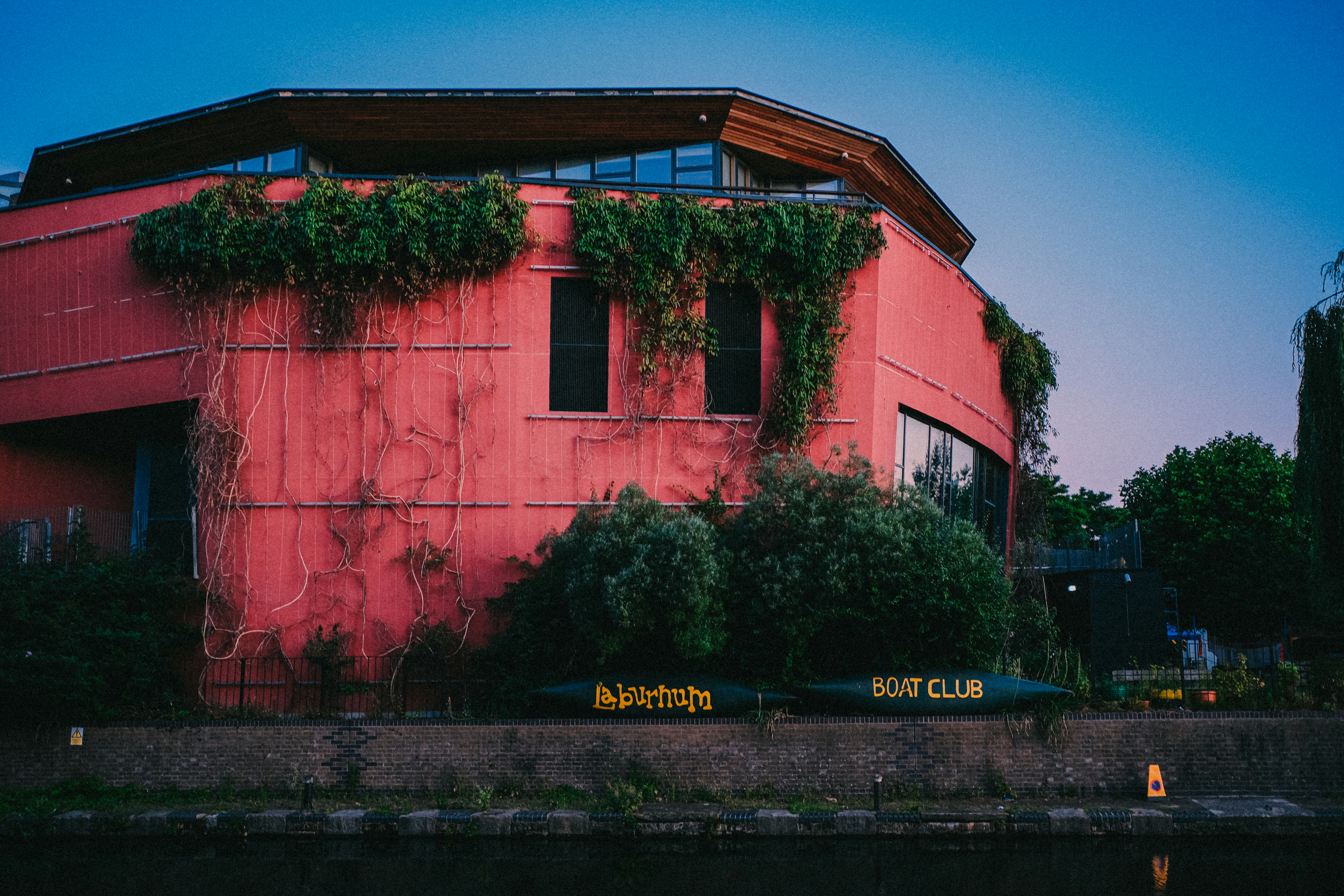 Modern building with vibrant pink walls and lush greenery by a canal at dusk.