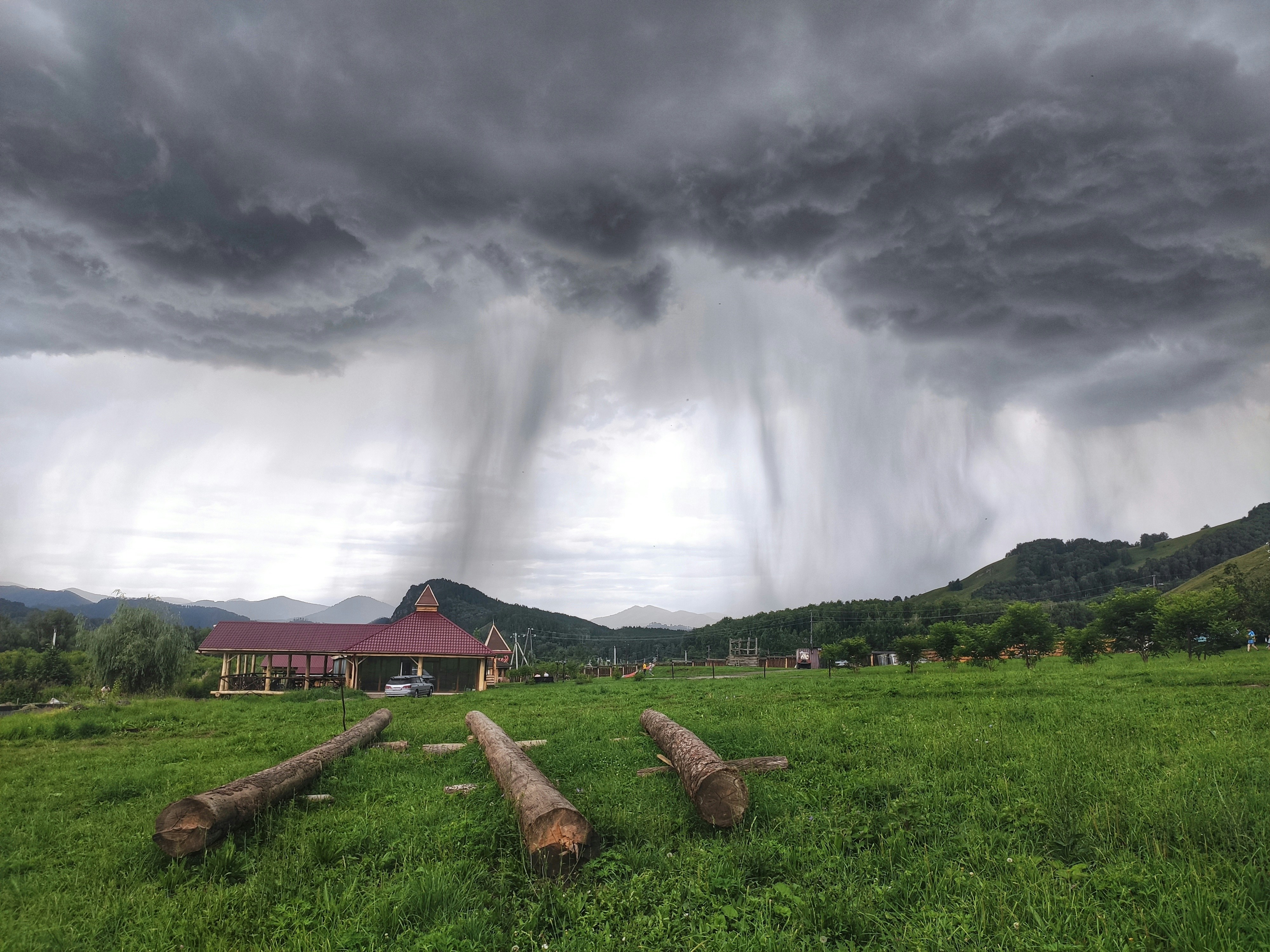 Logs on verdant grass under ominous clouds with rain descending near a mountain cabin.