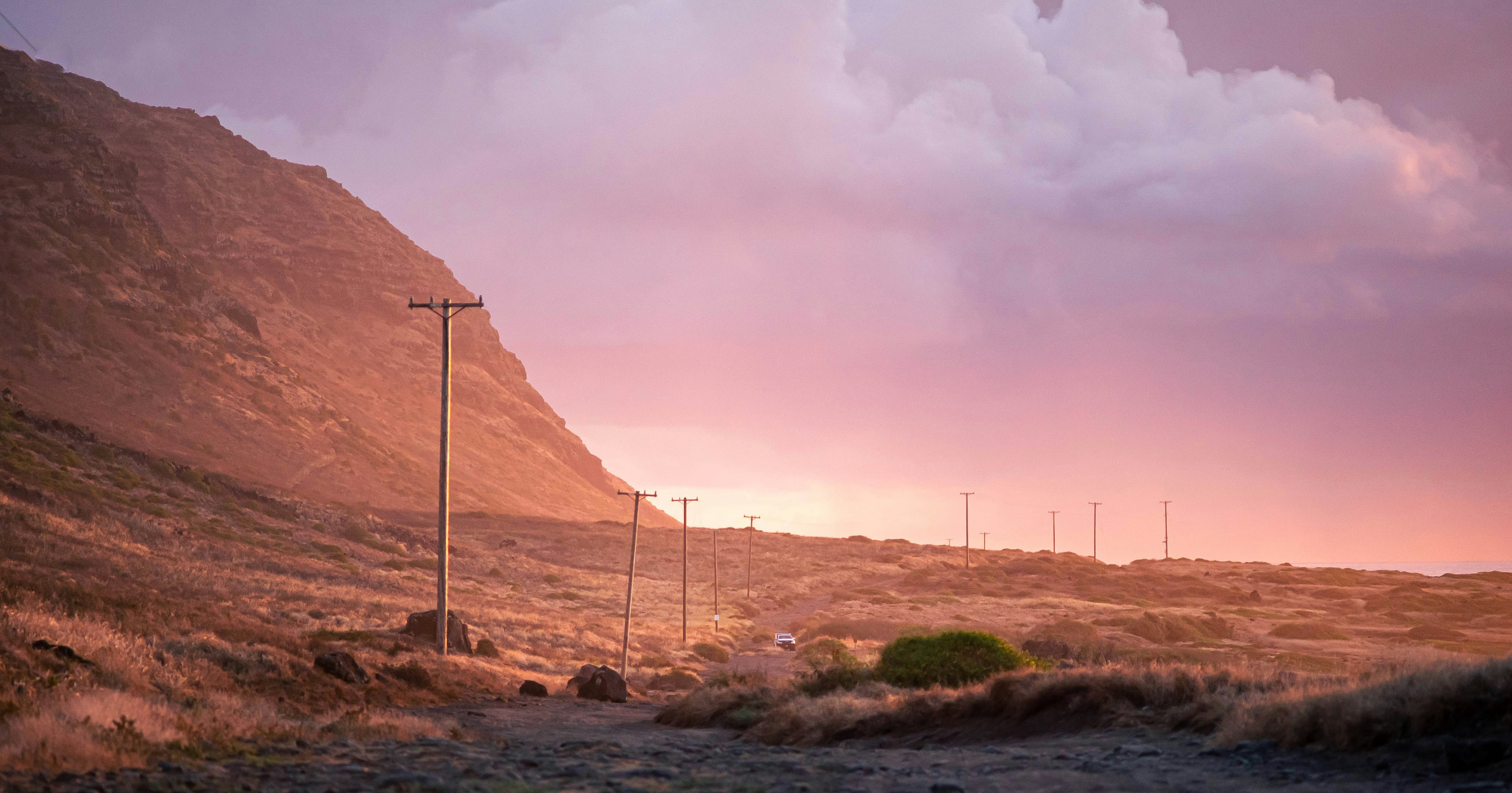 A single off-road vehicle makes its way back to the paved road along dirt during a Hawaii sunset, overlooked by telephone lines