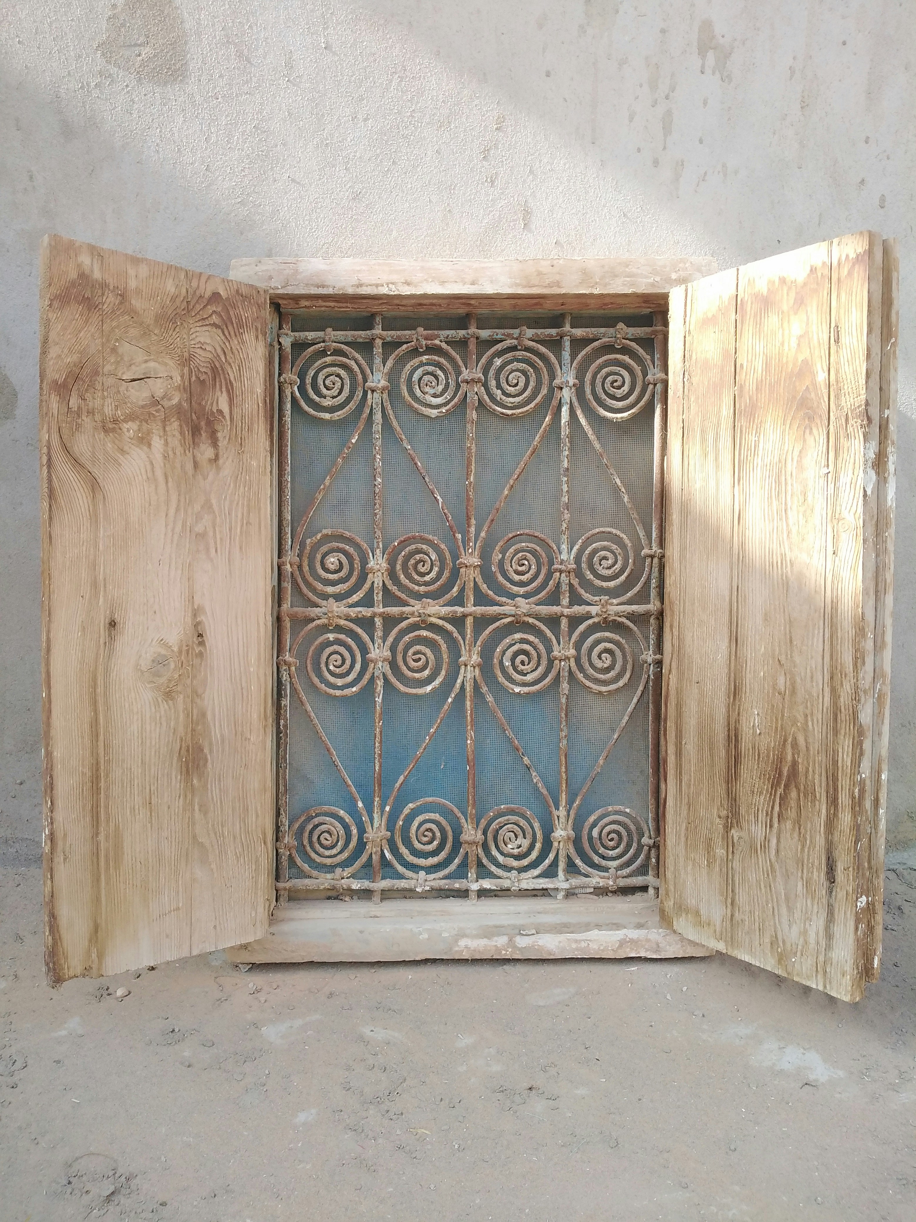 Sunlit Rustic Window: Weathered Shutters Framing a Turquoise Scrollwork Grille
