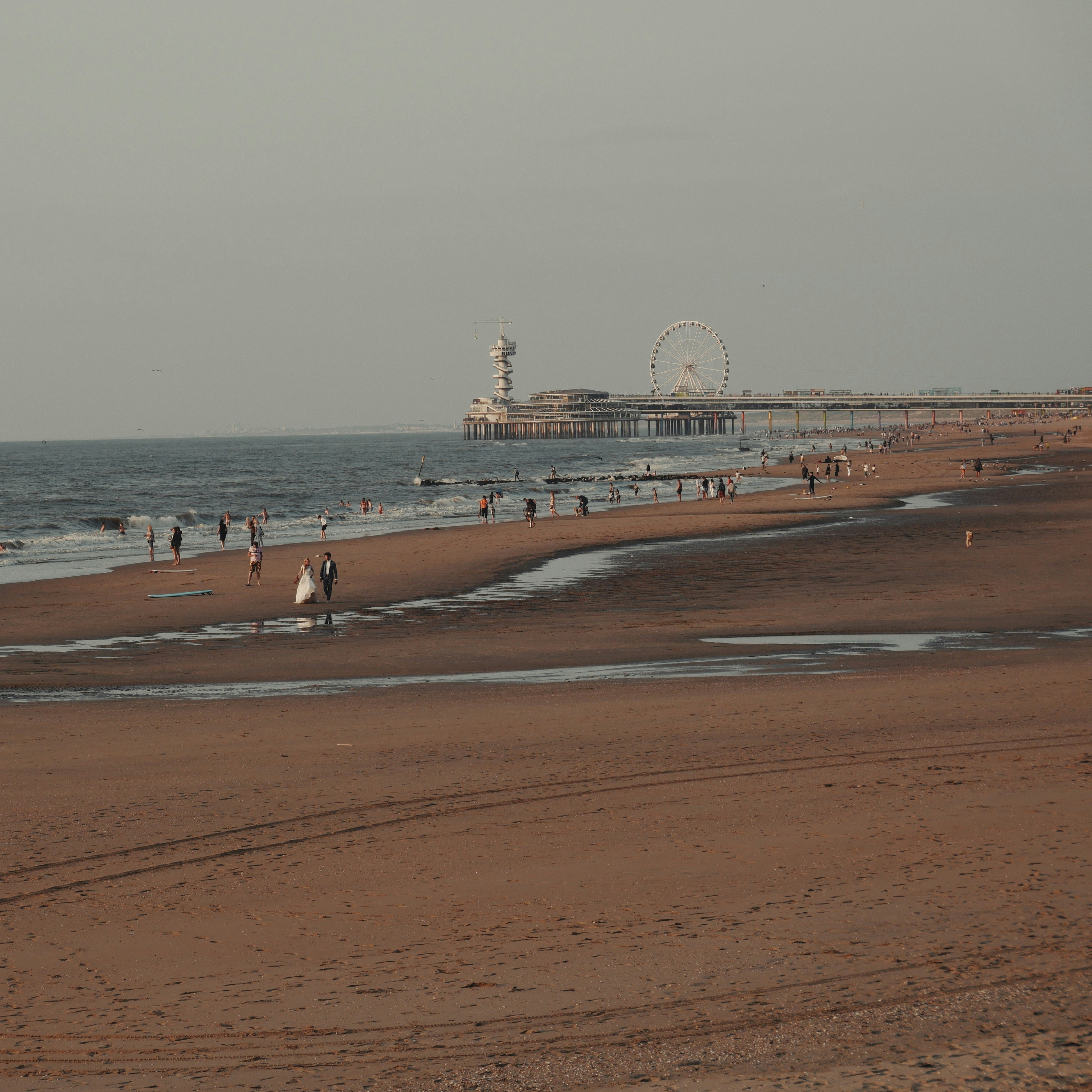 People walking along a sandy beach with gentle waves lapping at the shore, featuring a distant pier and a Ferris wheel.