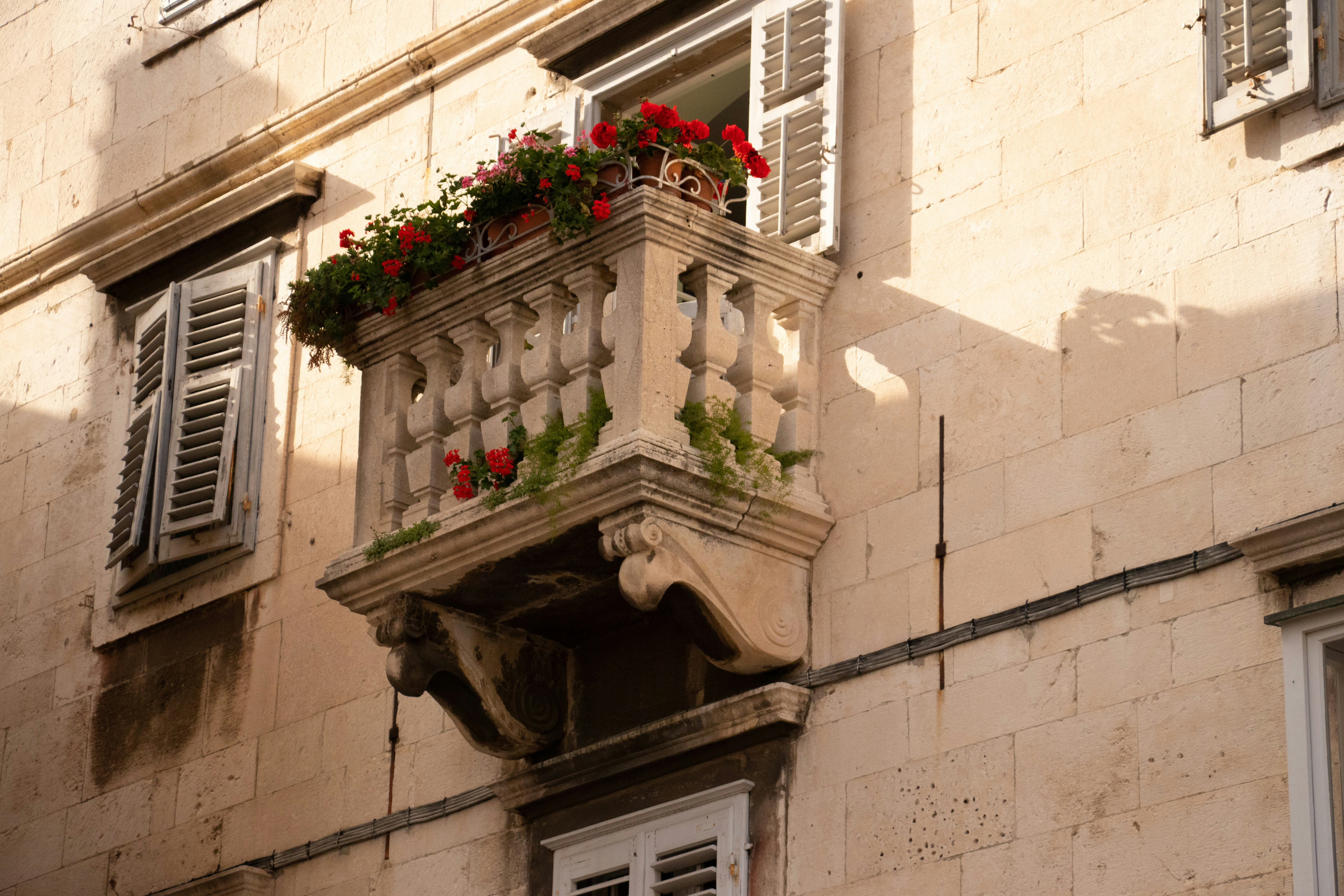 Ornate balcony adorned with vibrant flowers, set against a textured stone wall with shuttered windows. The scene reflects the charm of historic architecture.
