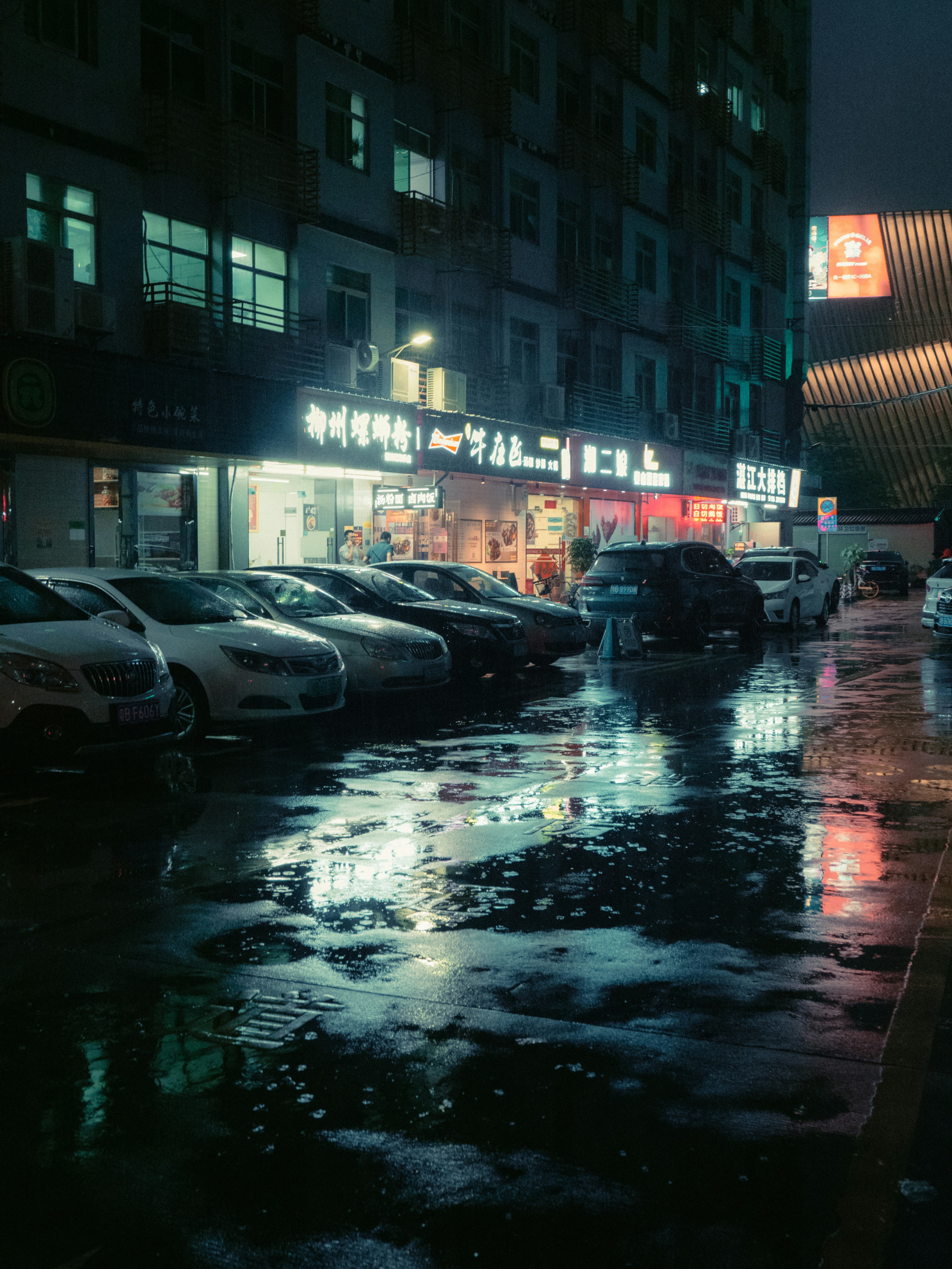 Wet street reflecting neon signs and parked cars in a bustling urban environment at night.