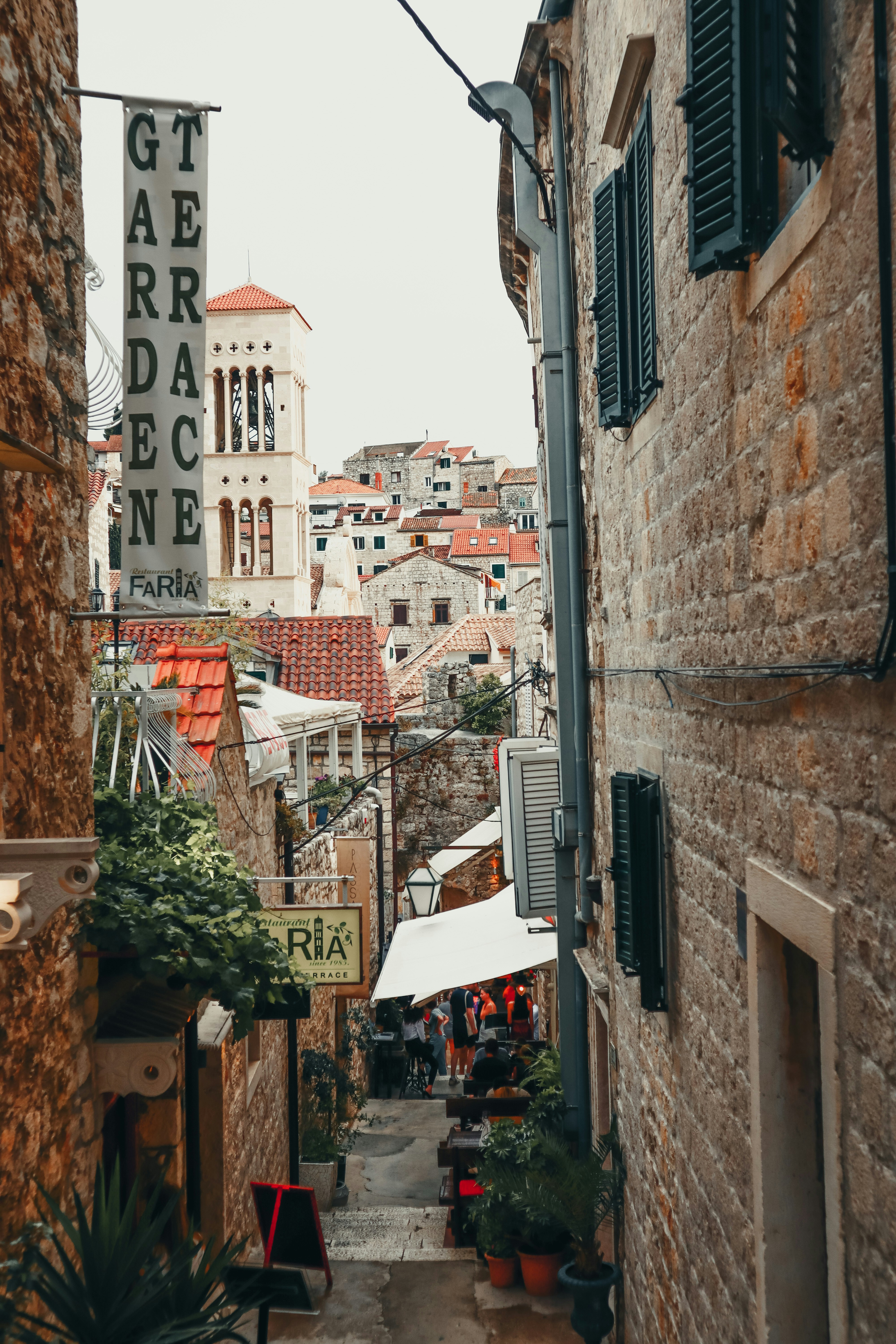 Historic stone alleyway lined with greenery and quaint signage, leading to a bustling terrace in the heart of the city.