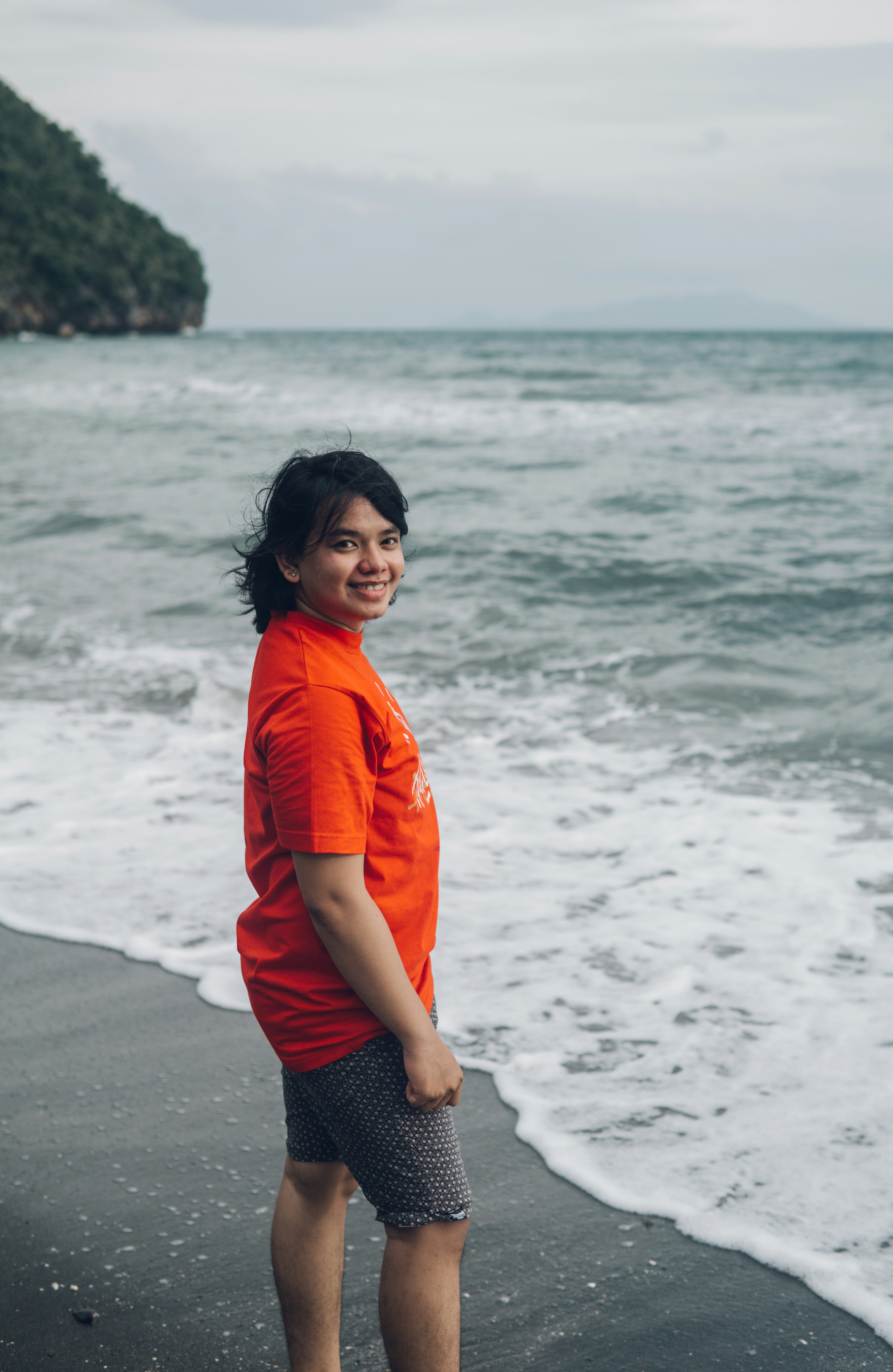 Person in a bright red shirt stands on a sandy beach near gentle ocean waves under a cloudy sky.