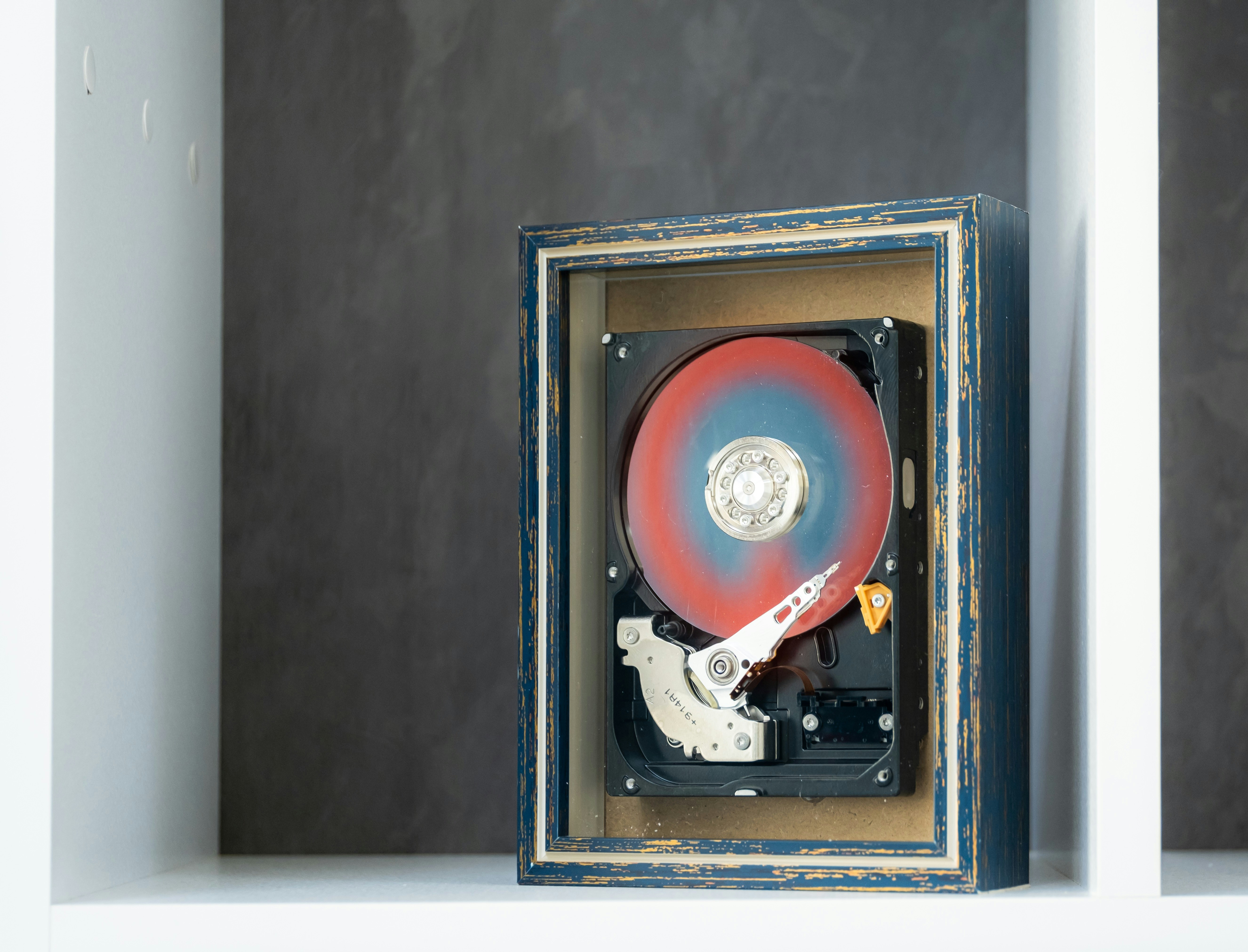 A vintage hard drive encased in a rustic frame, showcasing its intricate inner workings. The vibrant red and blue platter spins within a minimalistic white shelf backdrop.