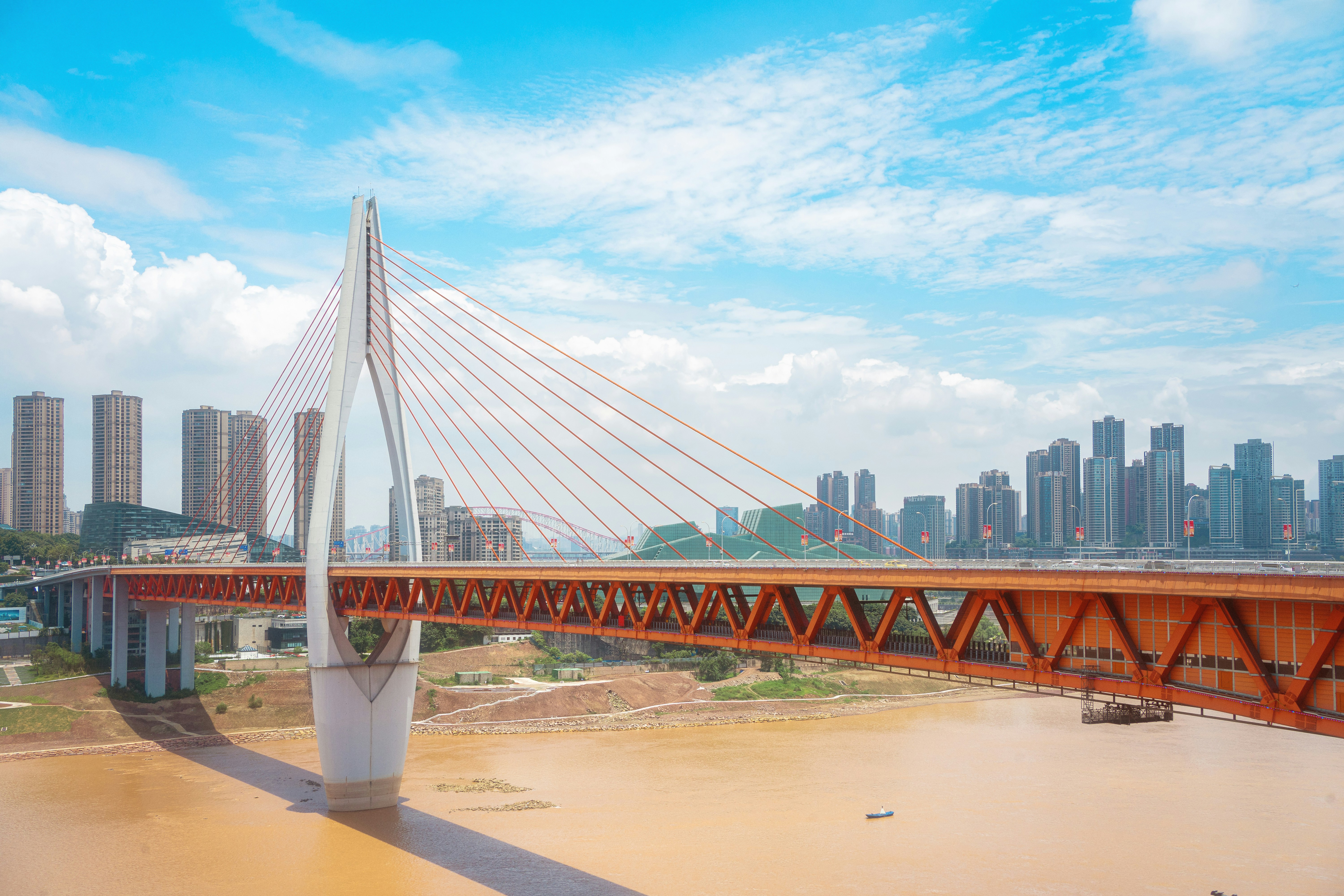 Suspension bridge with striking red beams extending over a wide river against a vibrant city skyline and blue sky.